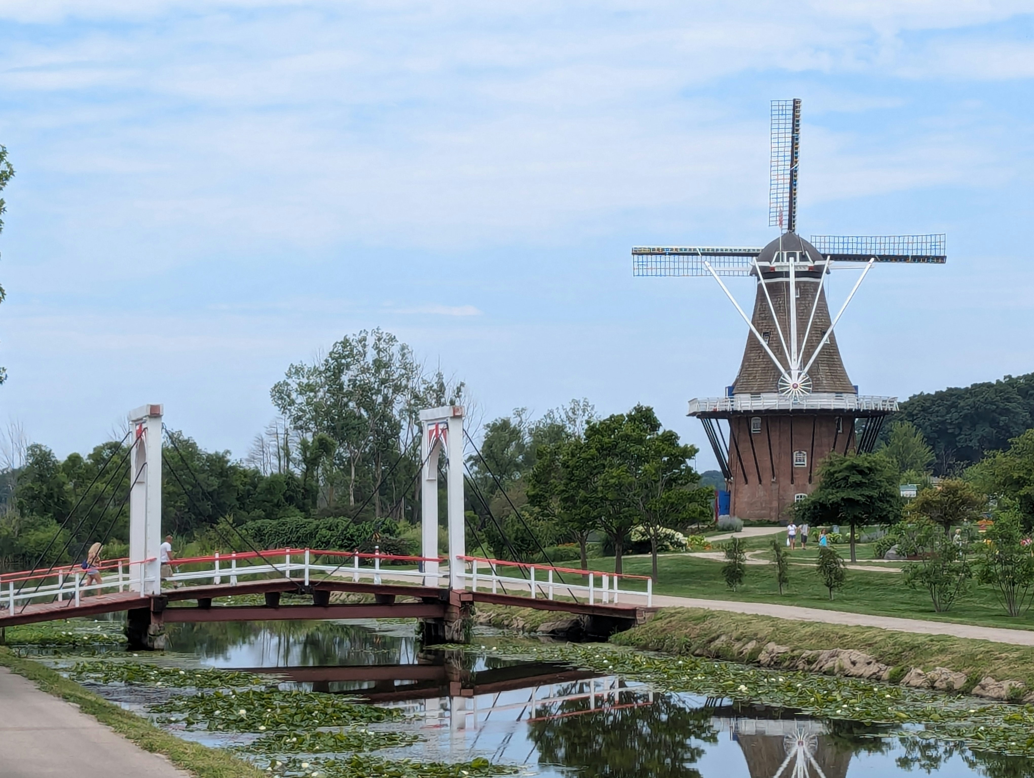 A charming windmill stands proudly beside a serene pond, framed by lush greenery and a quaint bridge. The scene captures a peaceful moment in nature.