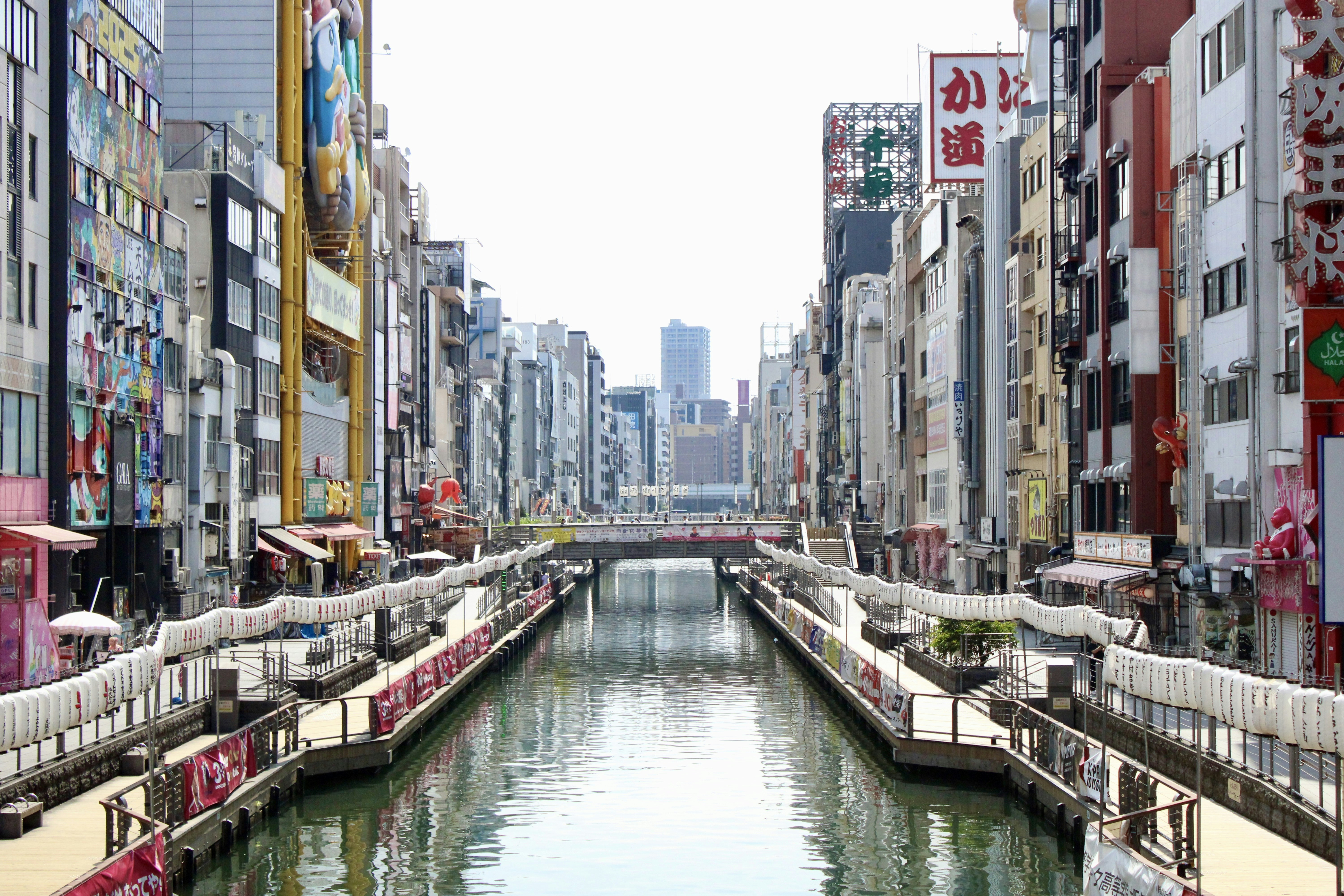 Vibrant cityscape featuring a canal flanked by bustling buildings and advertisements, showcasing urban life and reflection on water.