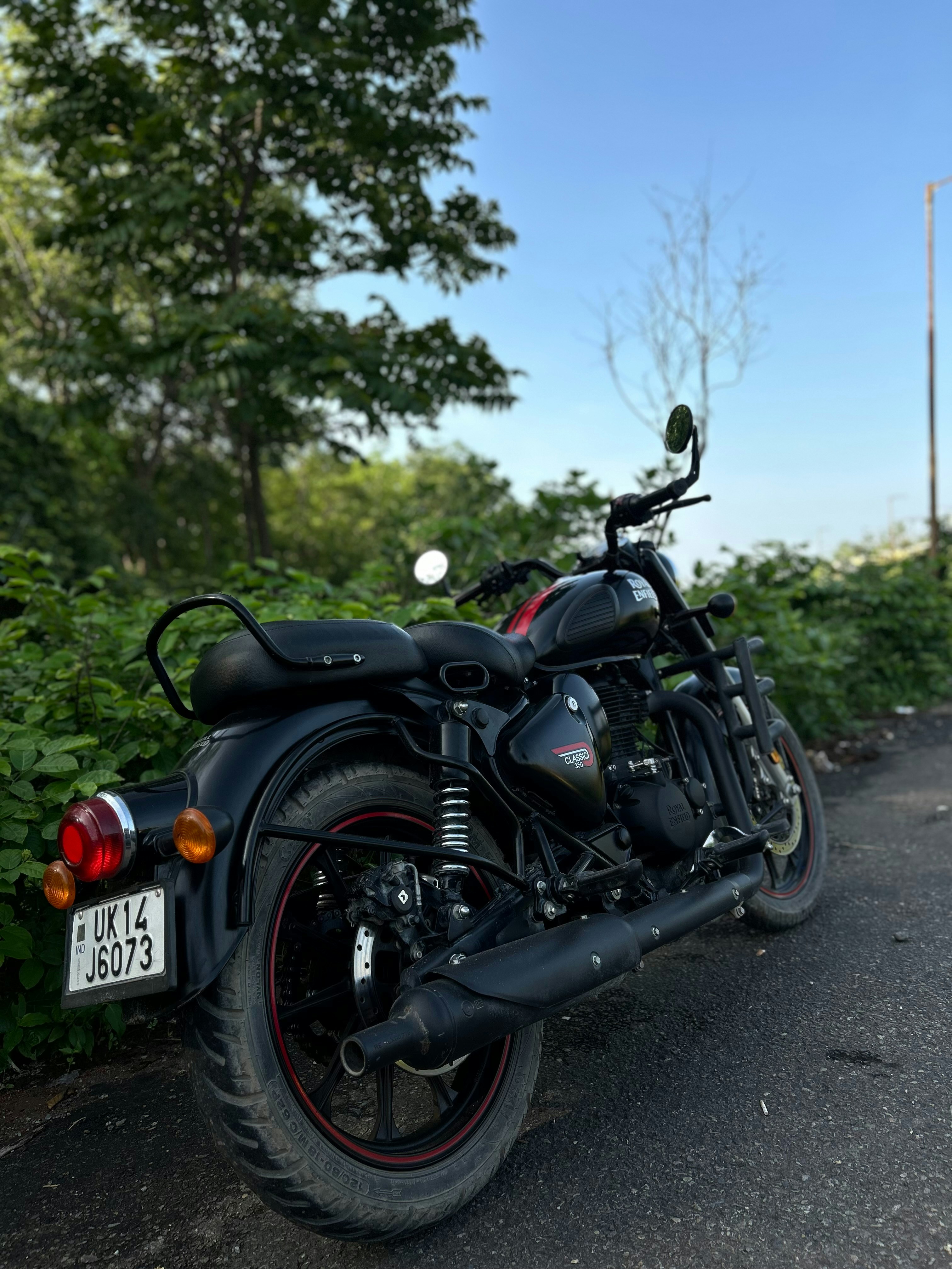 Black retro motorcycle with red accents parked along a paved roadside, framed by lush greenery and a clear blue sky.