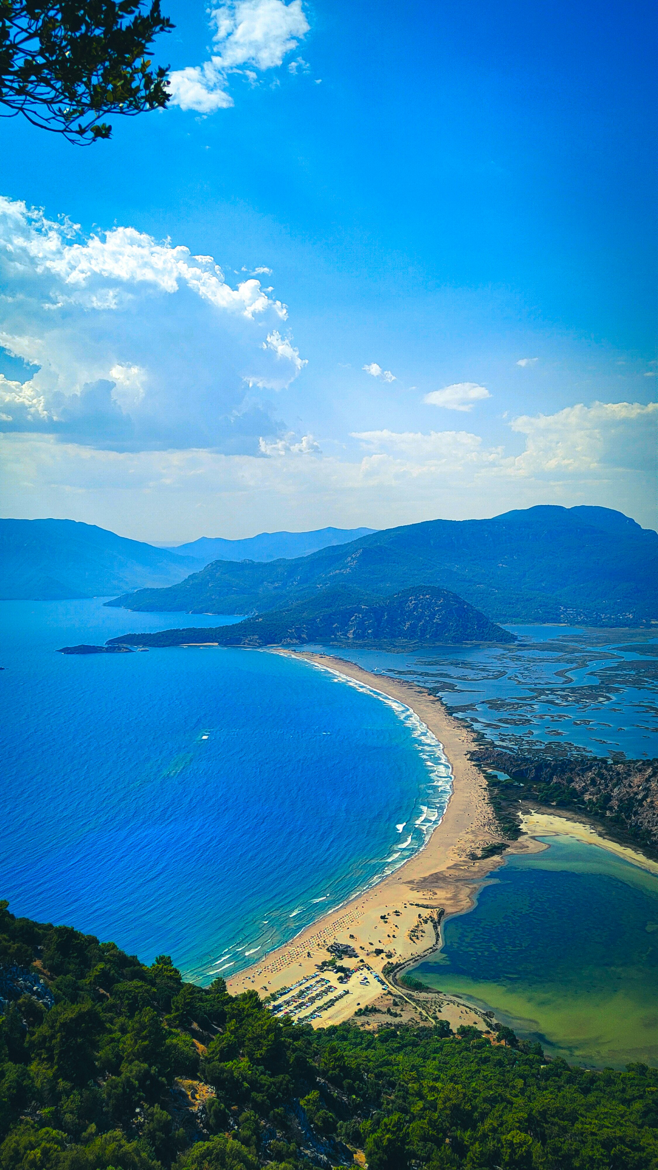 Aerial view of a sweeping beach curving along the coastline, framed by lush greenery and distant mountains. The vibrant blue waters contrast beautifully with the sandy shore.