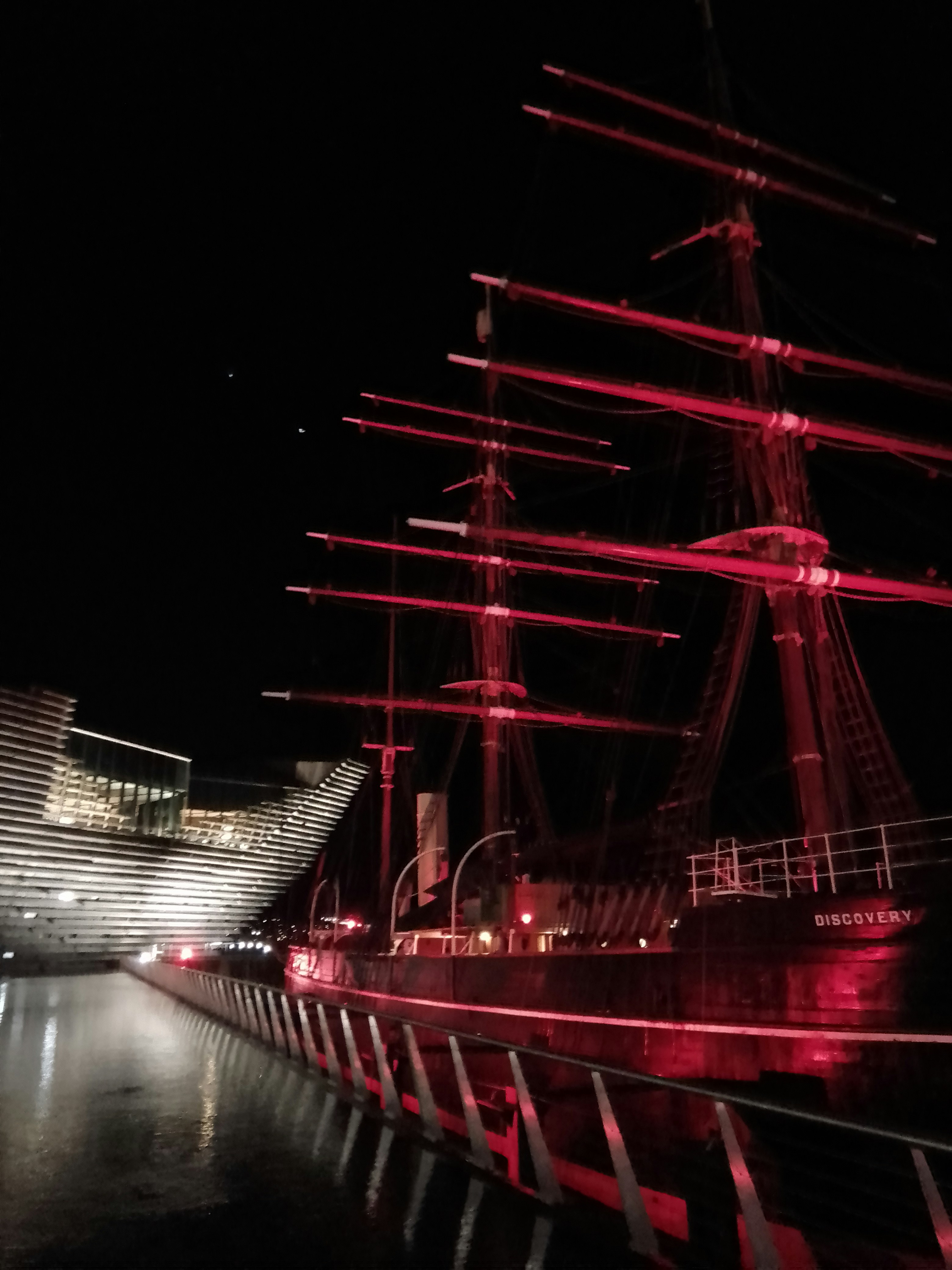Tall sailing ship, RSS Discovery, illuminated in red, in dry dock in front of floodlit V&A Museum.