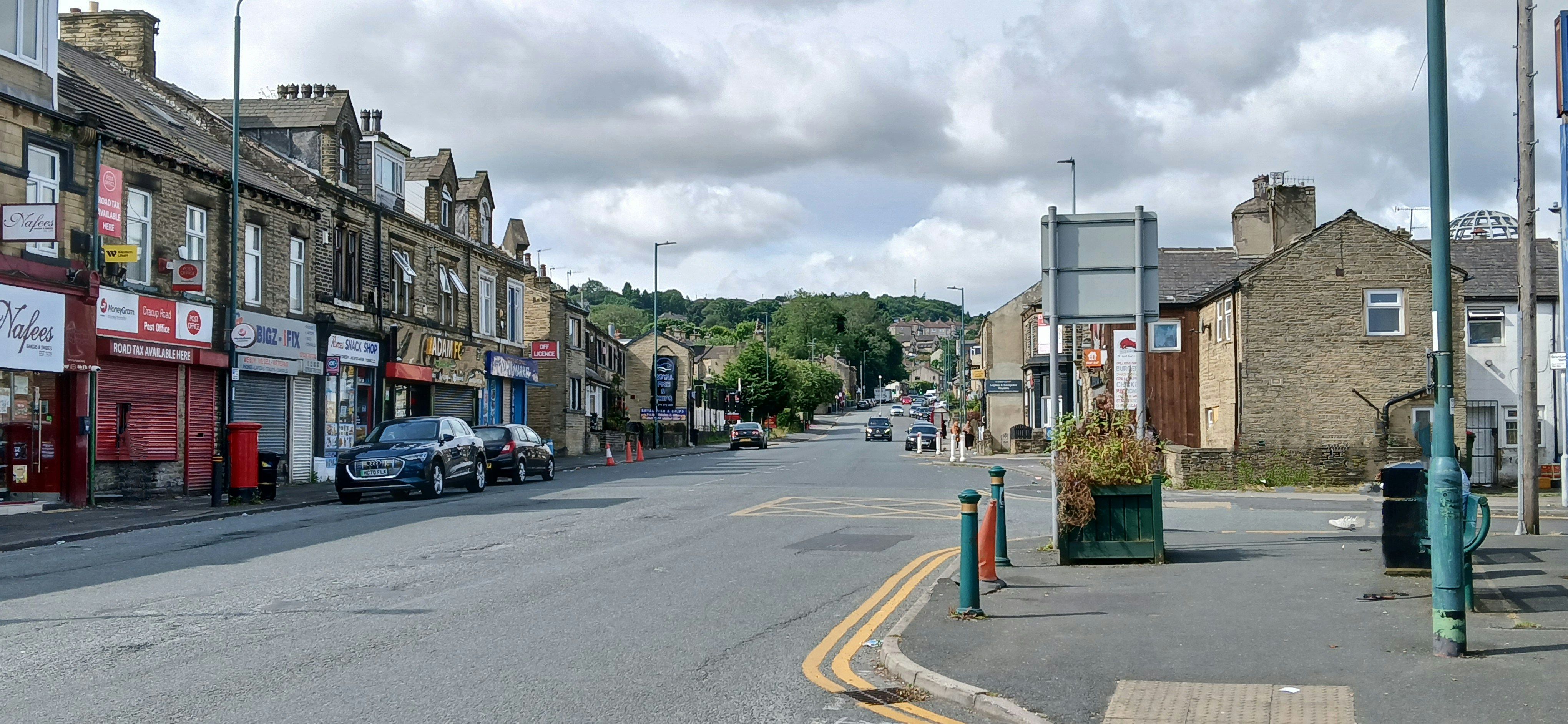 Quiet street scene in Great Horton with shops lining the road under a cloudy sky.