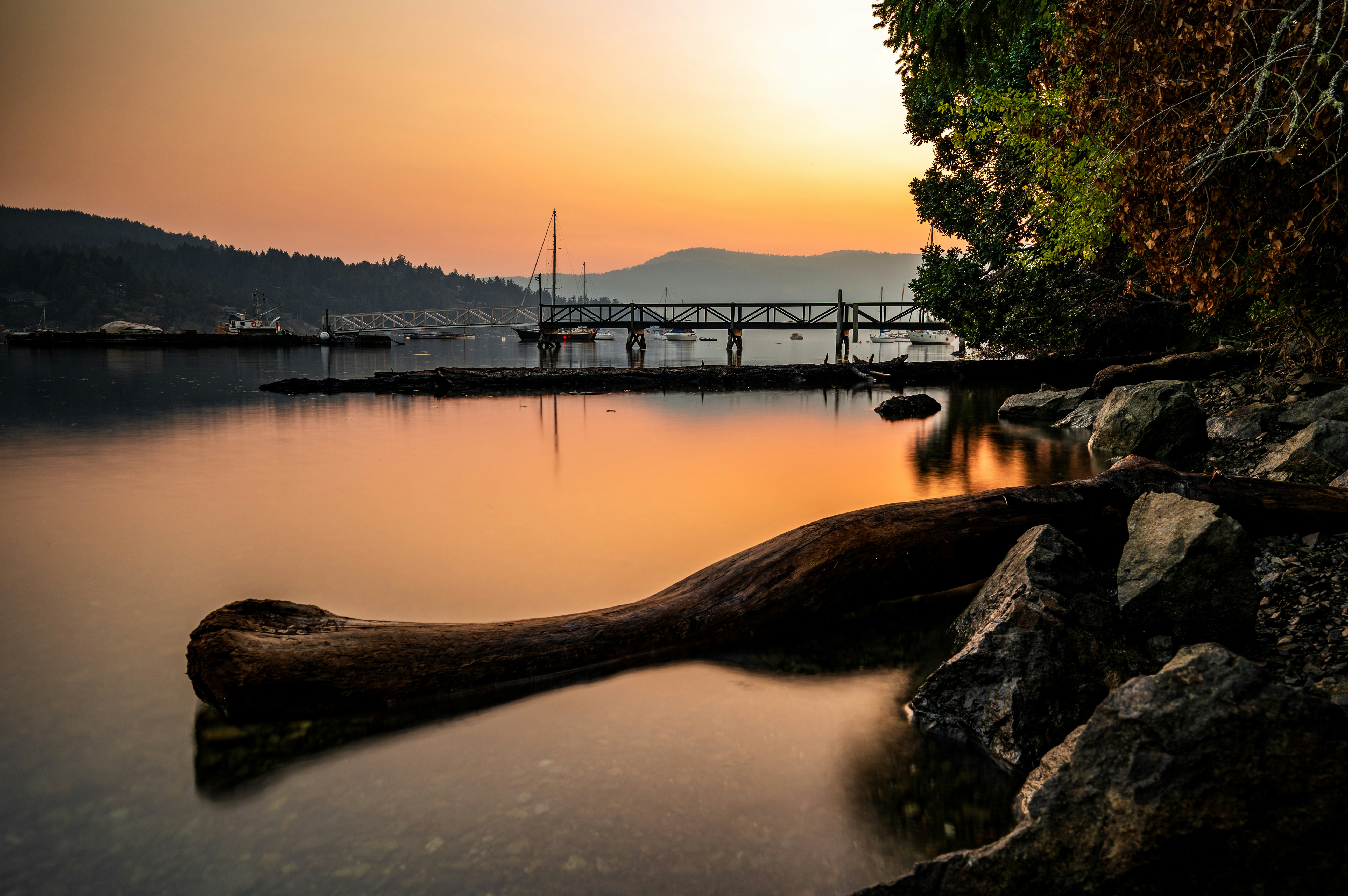 Calm lake reflecting a vibrant sunset with rocks and a silhouetted tree in the foreground.