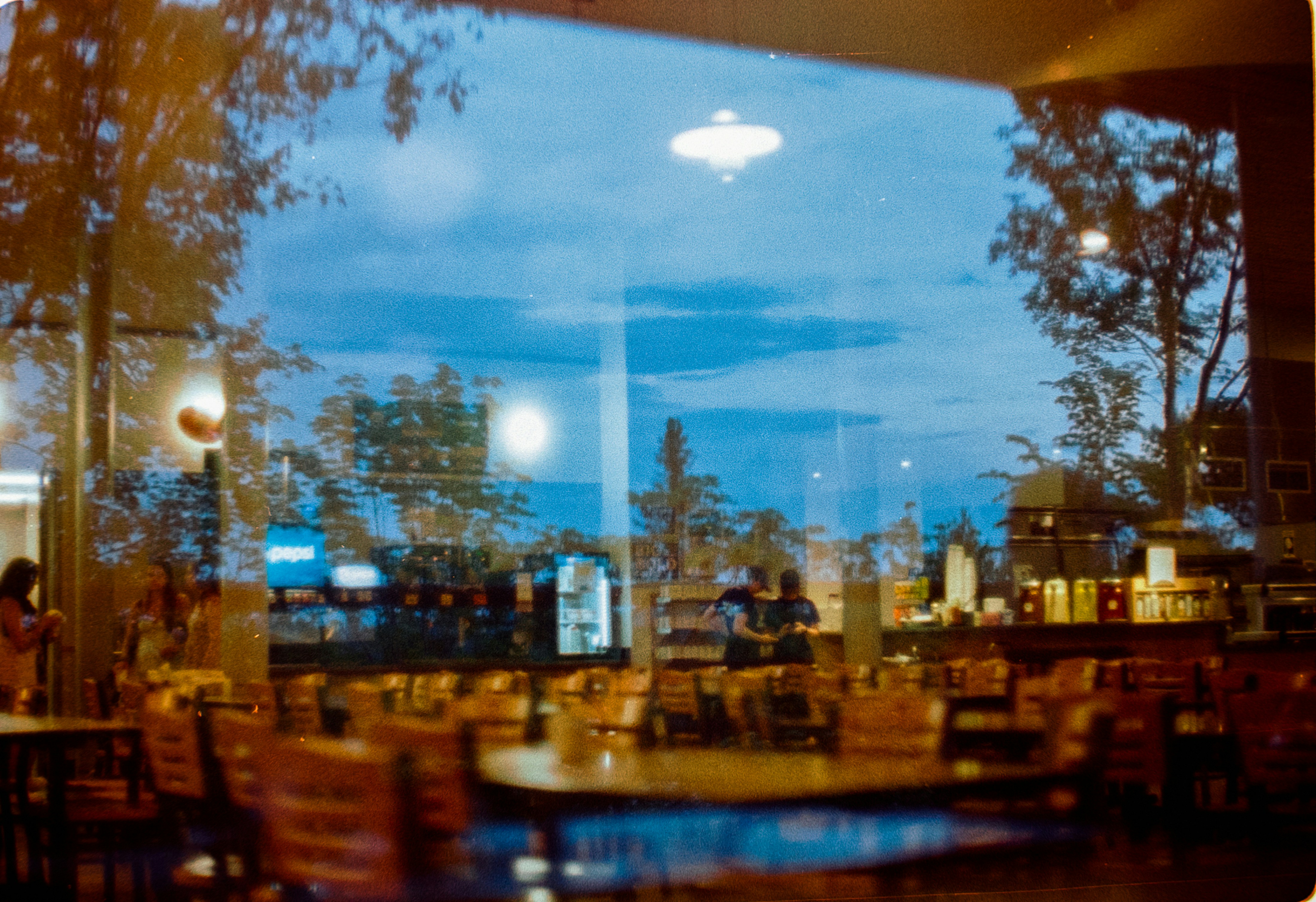 Photograph of a cafe interior seen through a glass window. Reflections of tables and chairs blur the scene while a blue sky and trees appear outside.