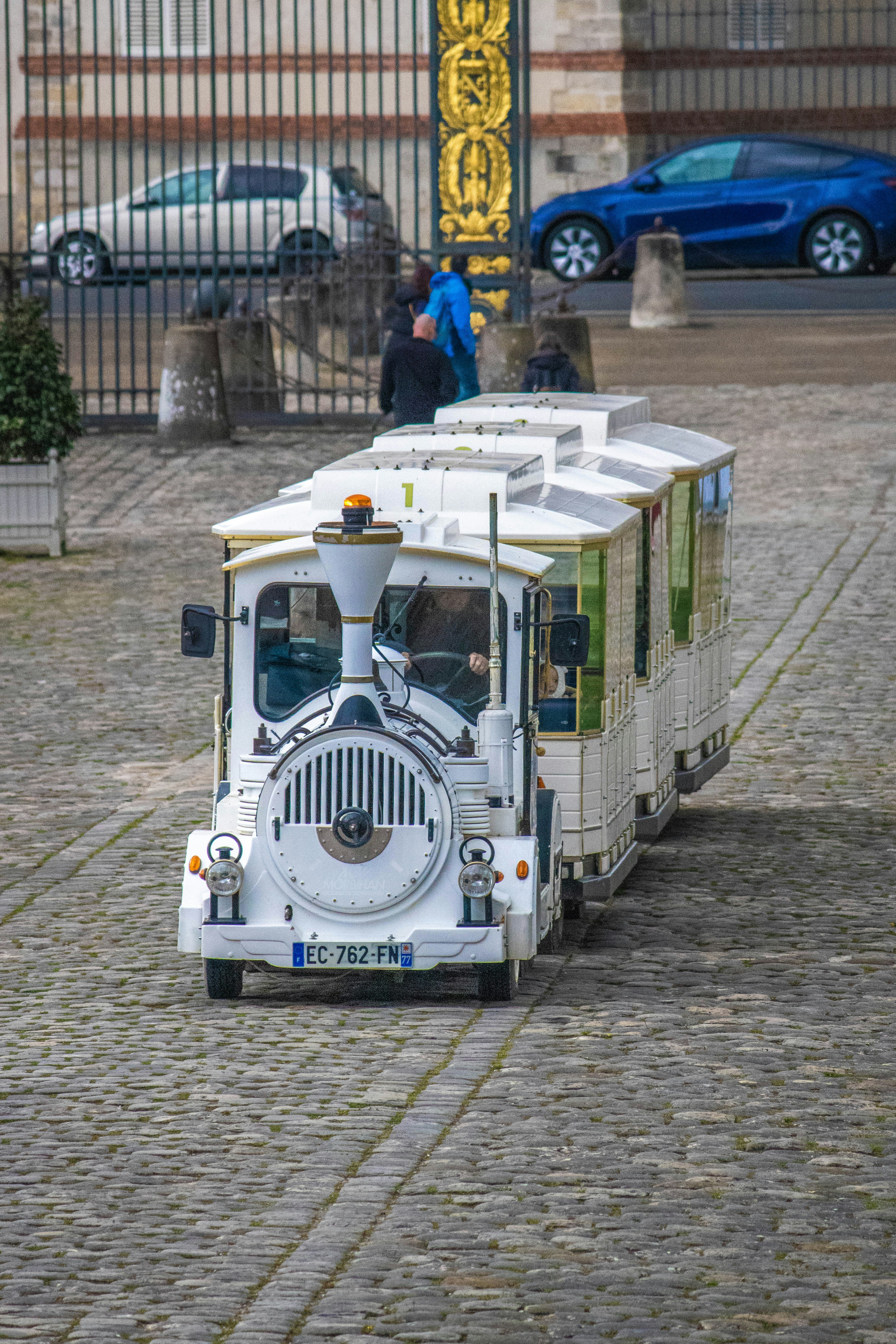 White Miniature Steam Locomotive Rounds a Cobblestone Lane by an Ornate Gate