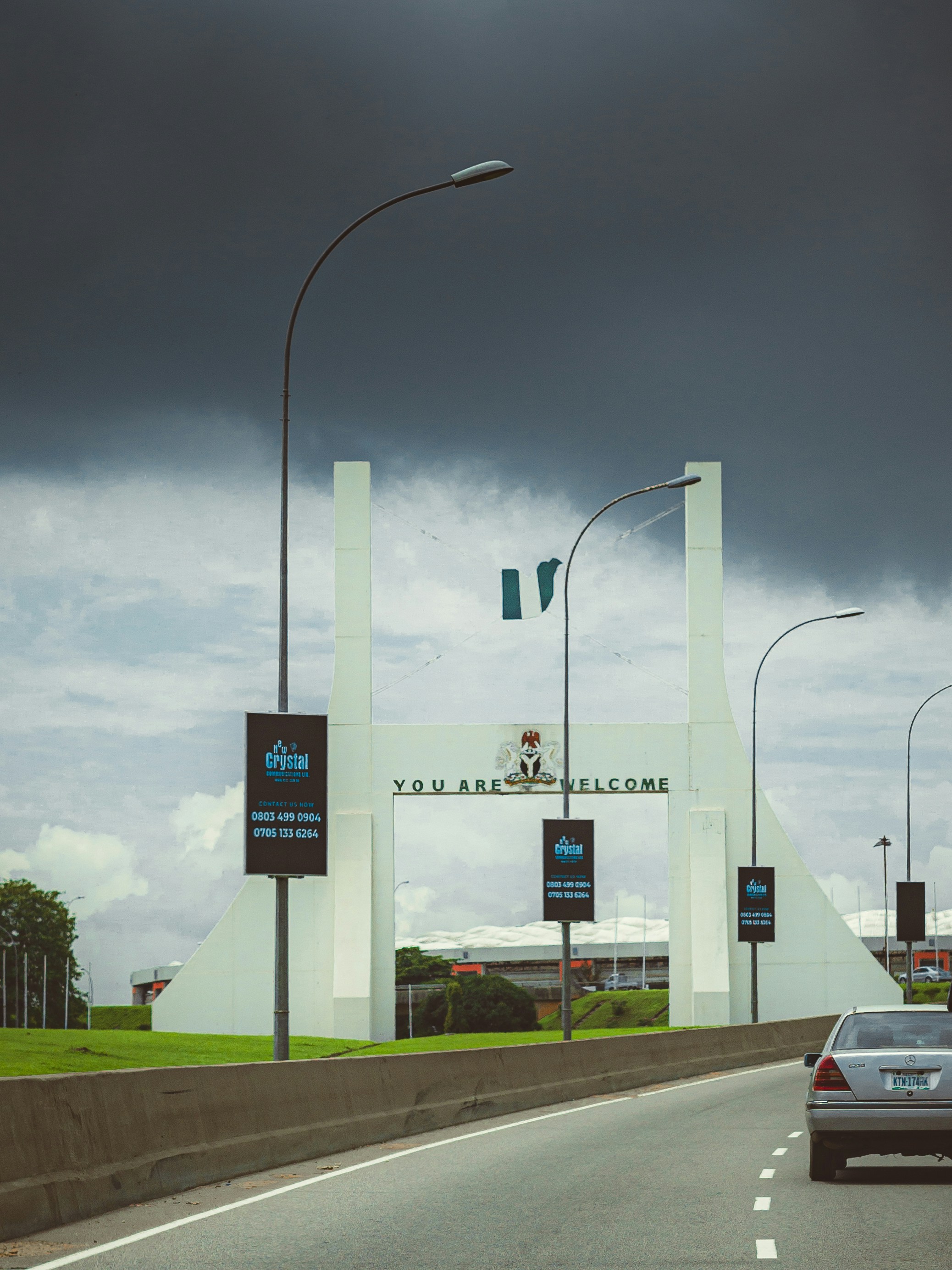 Welcome sign adorned with the national flag, flanked by advertisements, set against a moody sky.