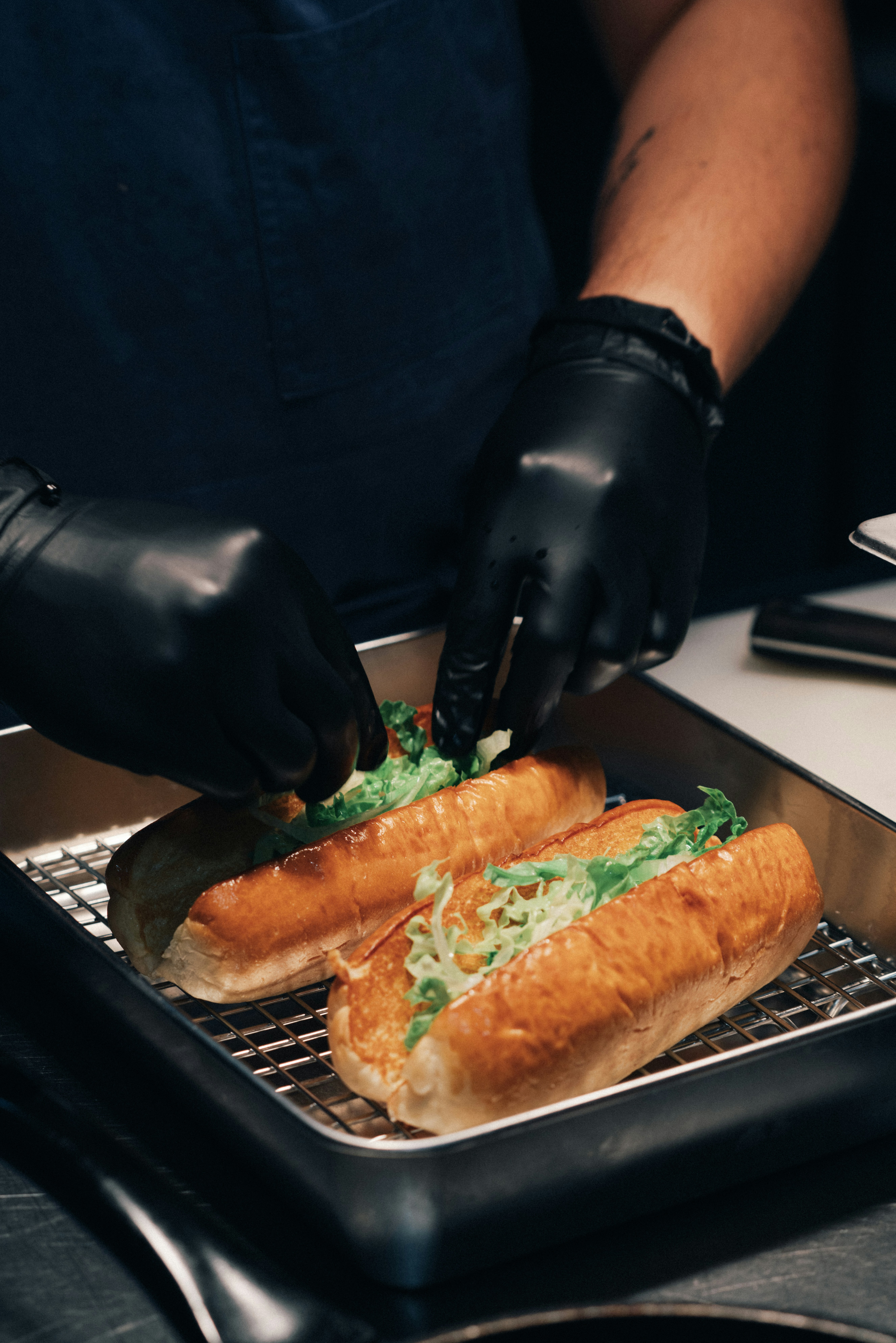 Gloved Craft: Precision lettuce tuck on two hot dogs in a stainless prep tray