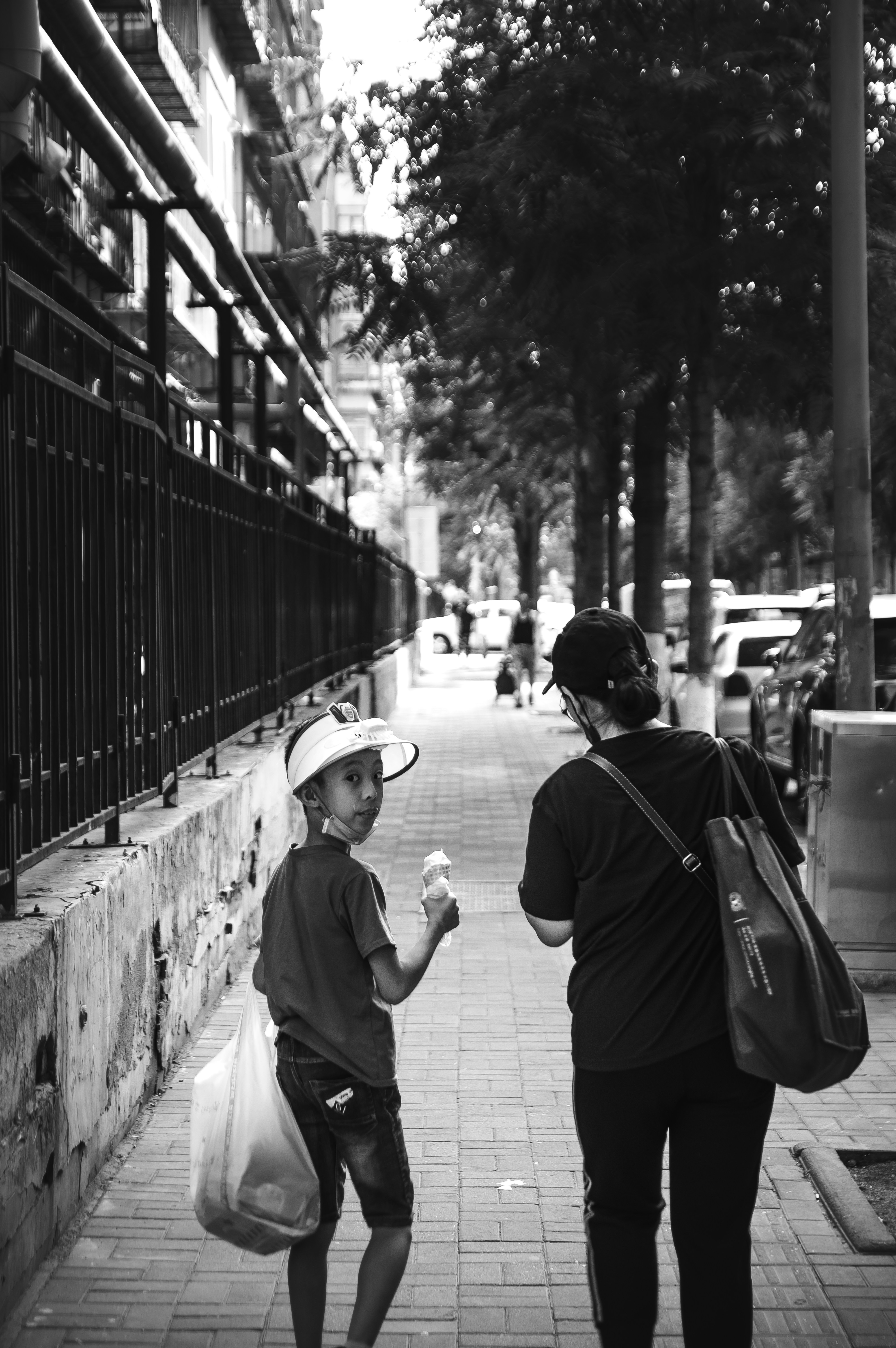 A black-and-white street photograph captures a boy in a hat with a bag and ice cream glancing back toward the camera as an adult walks ahead along a tree-lined city sidewalk.