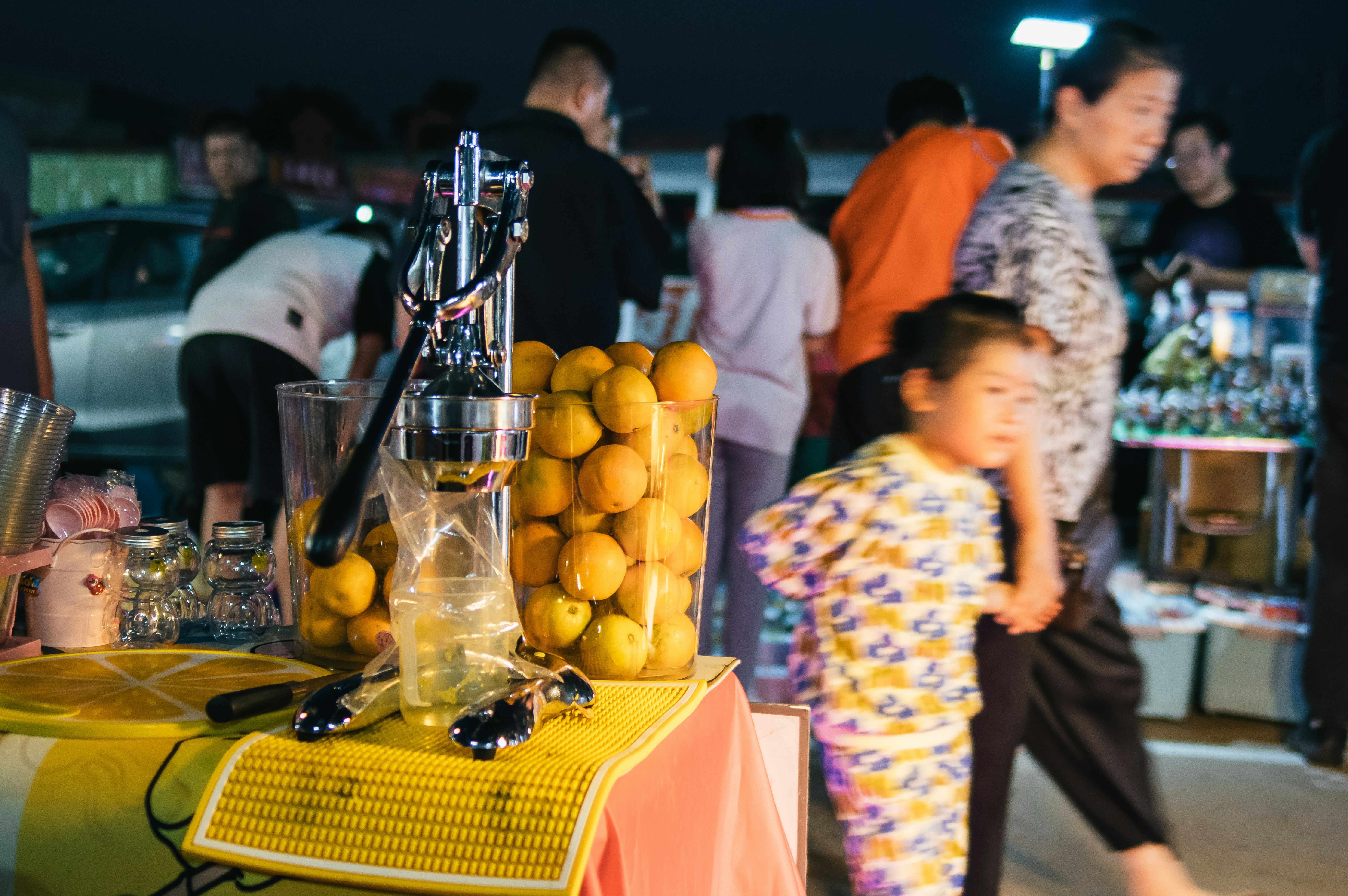 Night market scene centered on a lemon juice press atop a yellow table with bright lemons. Figures blur in the busy background, conveying lively evening activity.