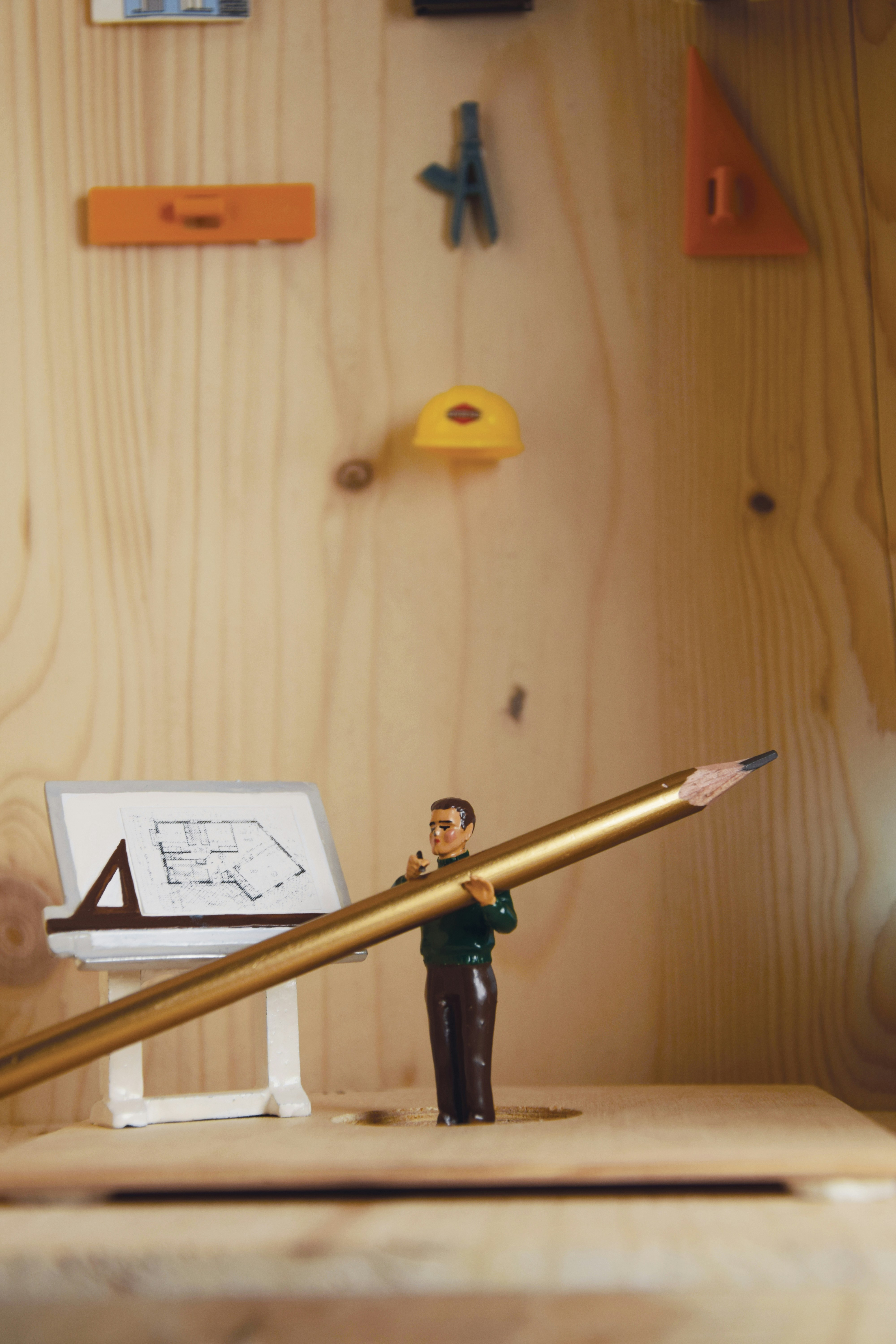 A miniature architect holding a big pencil stands next to a miniature architect's table, with miniature architect tools hanging on the wooden wall beside him.
