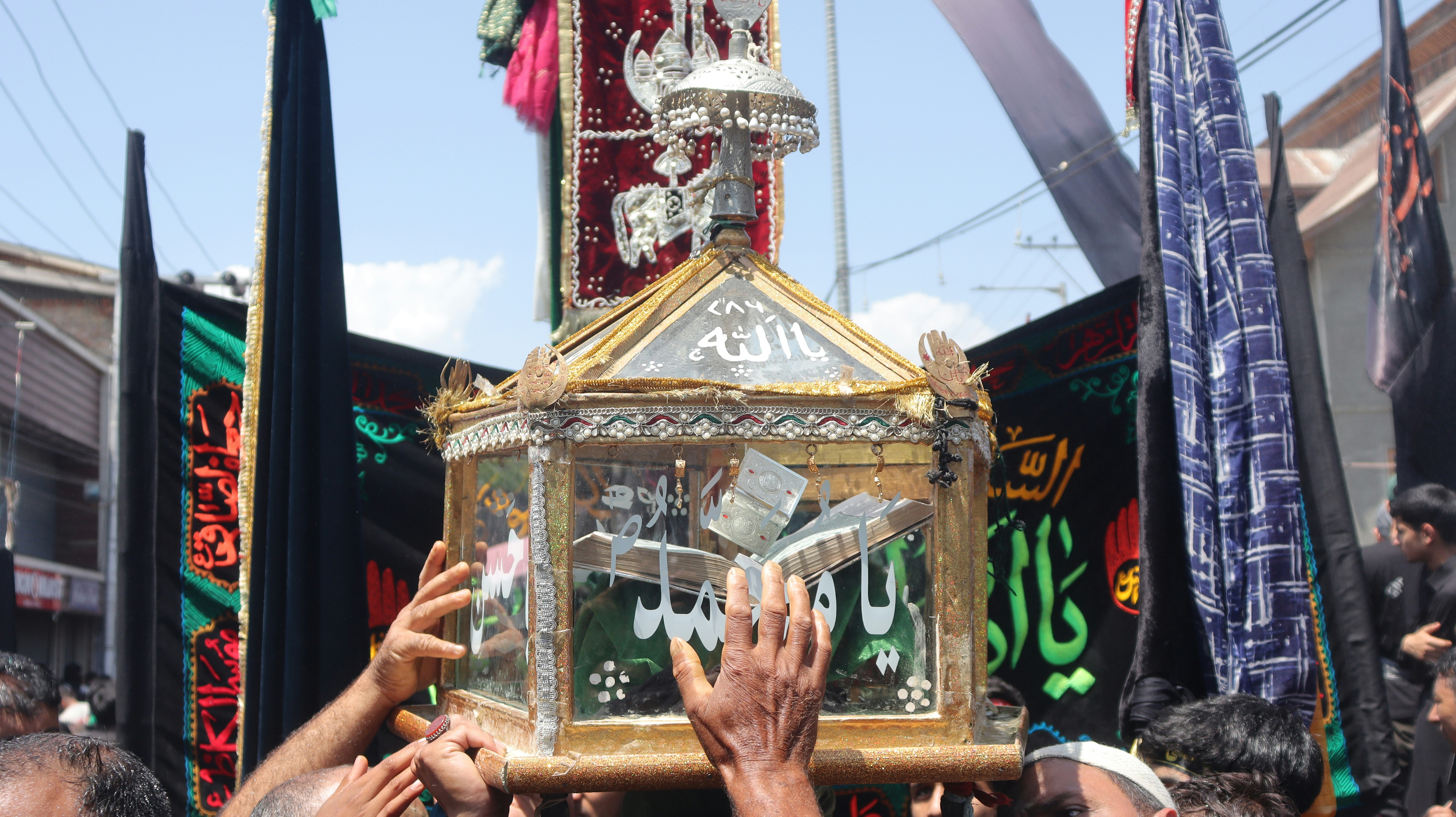 Participants carry a beautifully adorned container during a religious procession, surrounded by vibrant banners and flags. The atmosphere is charged with devotion.