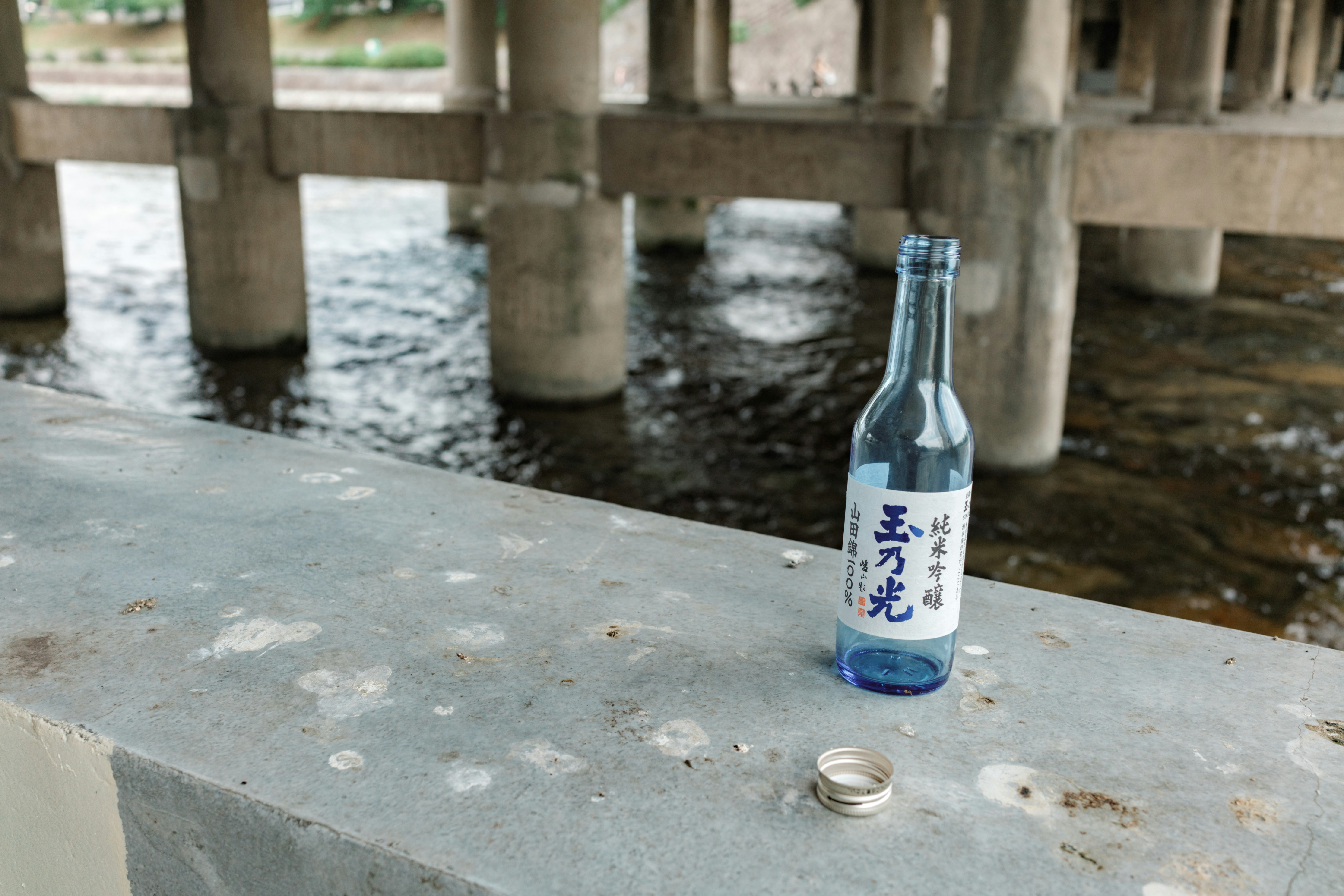 Blue glass bottle with a white label sits on a weathered concrete ledge beside a river under a concrete pier; the cap rests nearby.