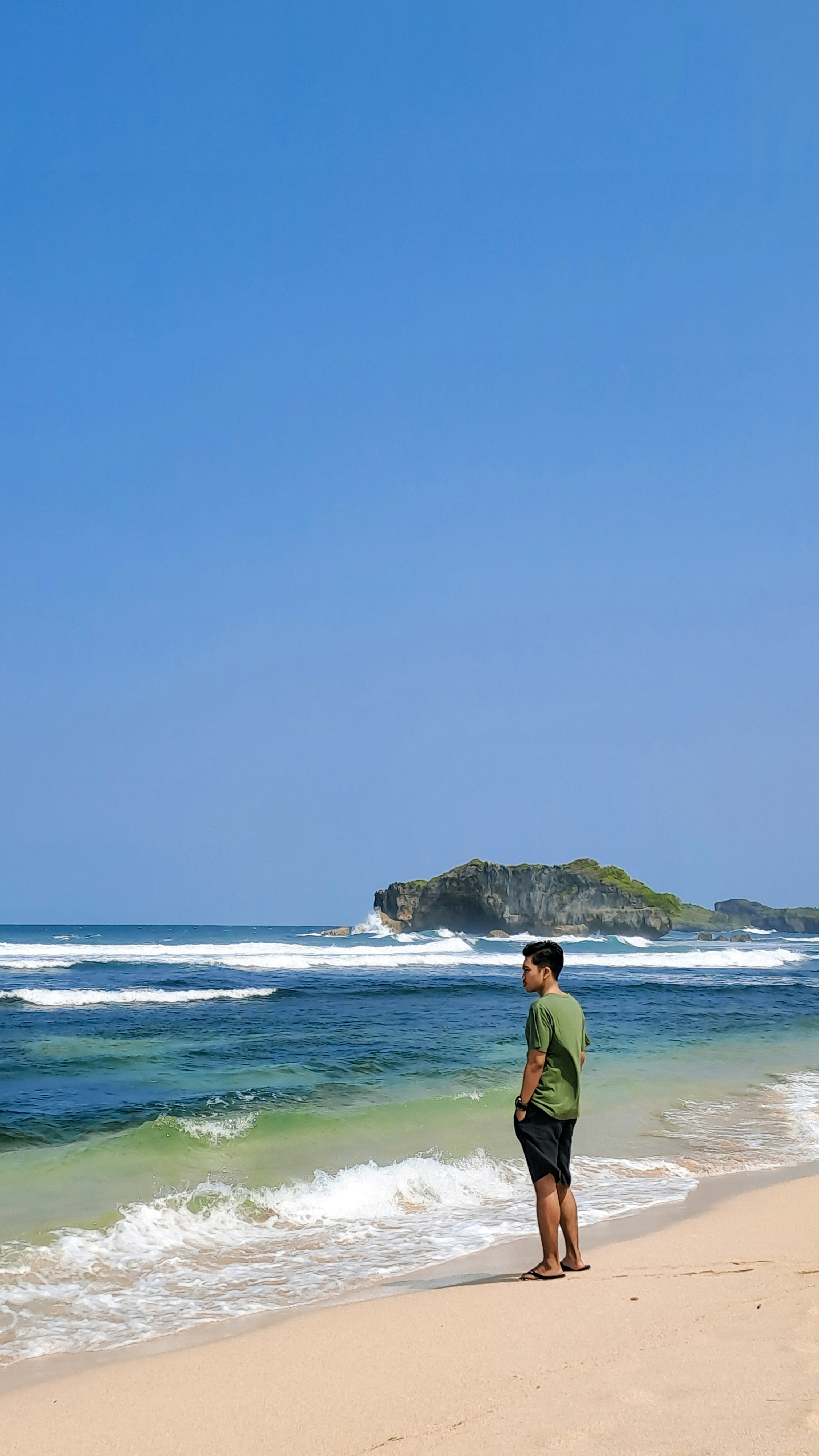 A man in a green shirt stands at the water's edge, facing the sea as waves roll in toward a distant rocky islet.