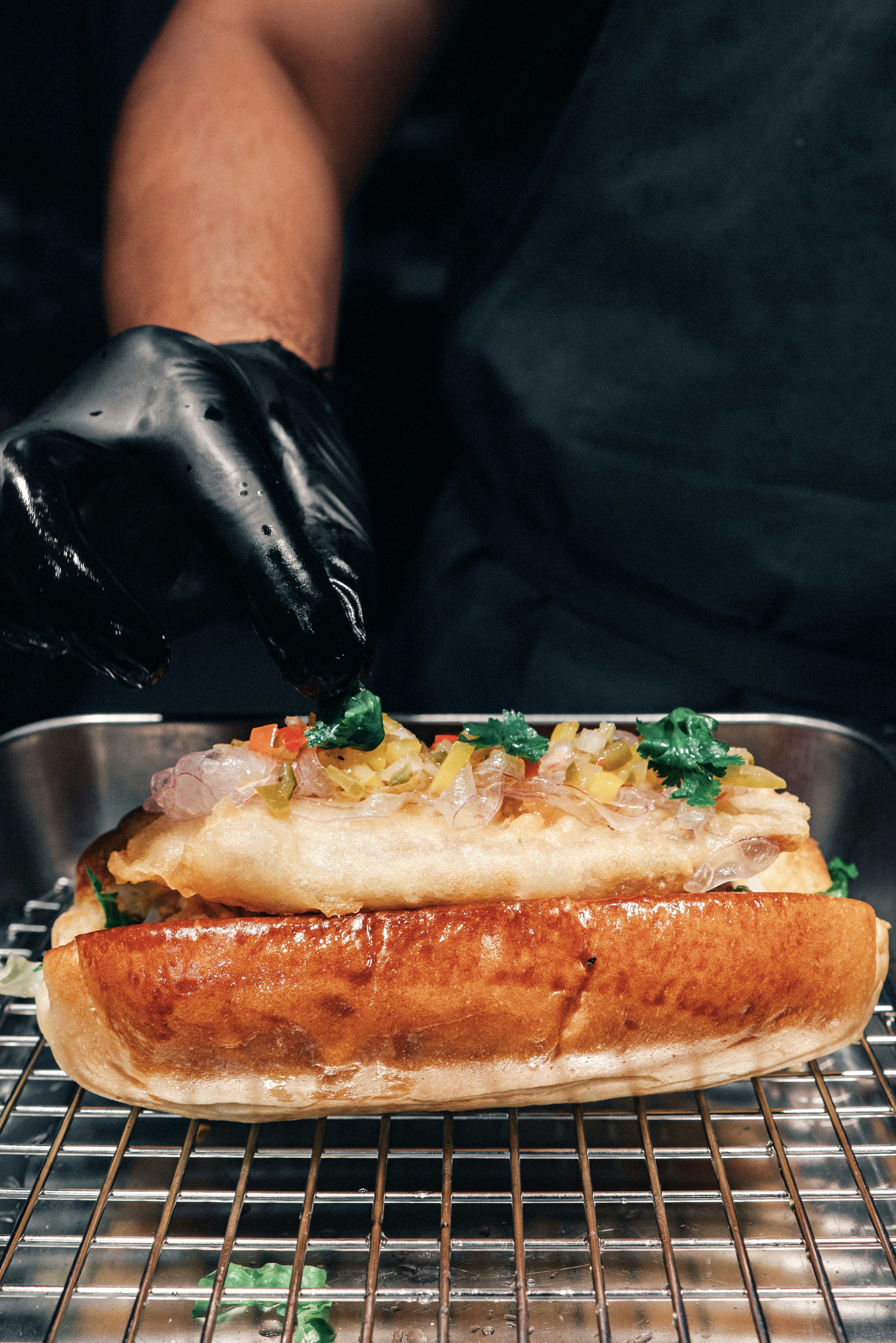 Savory hot sub resting on a rack, topped with chopped onions and herbs; a gloved hand applies finishing touches. A food preparation scene featuring a glossy, toasted bun and metallic tray.