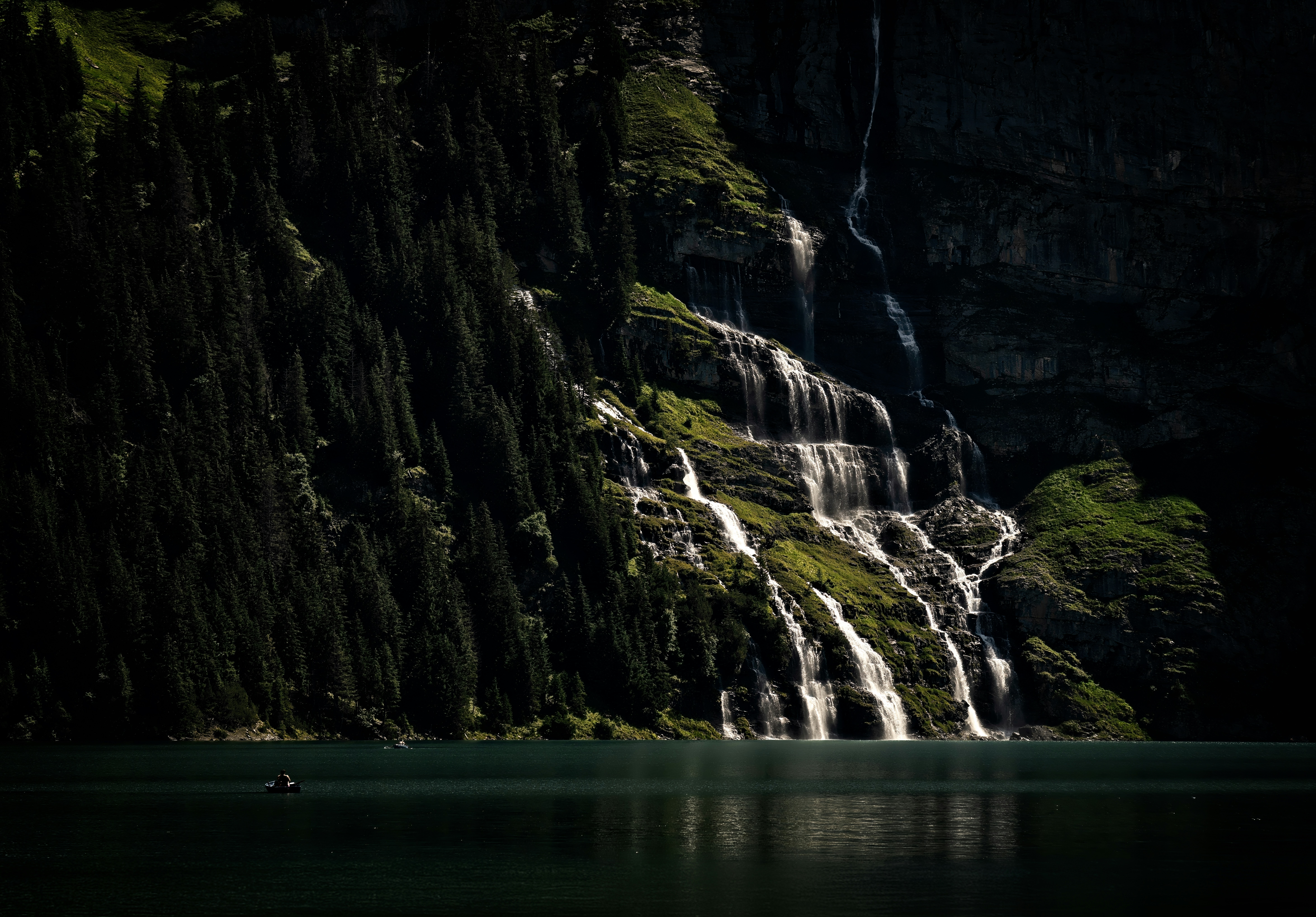 Waterfall cascading down lush green cliffs into a serene lake under dappled sunlight.