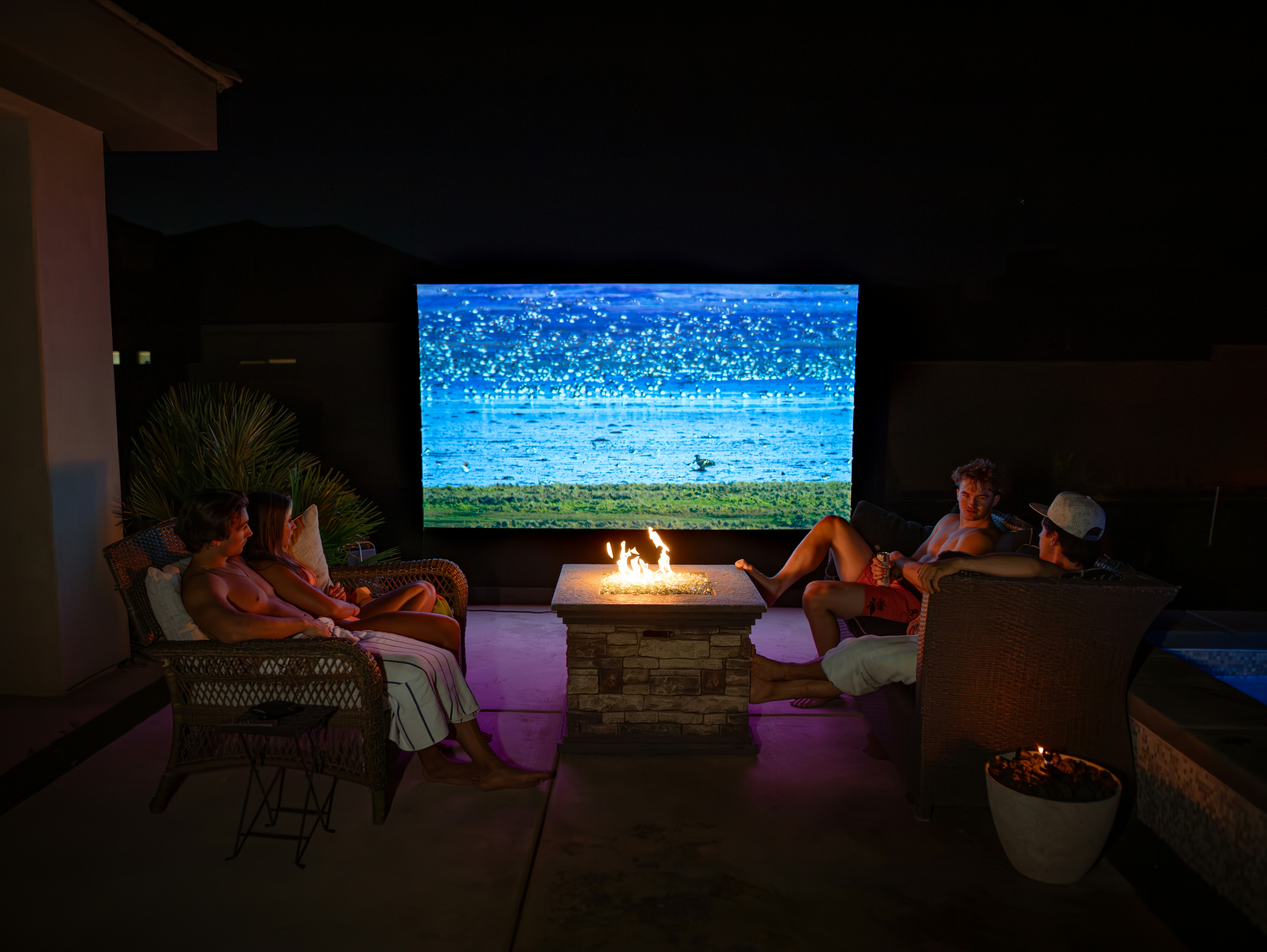 A group of people gathered around a fire pit, enjoying an immersive viewing experience with the AWOL Vision 4K UST Laser Projector. | A group of people sitting around a fire pit