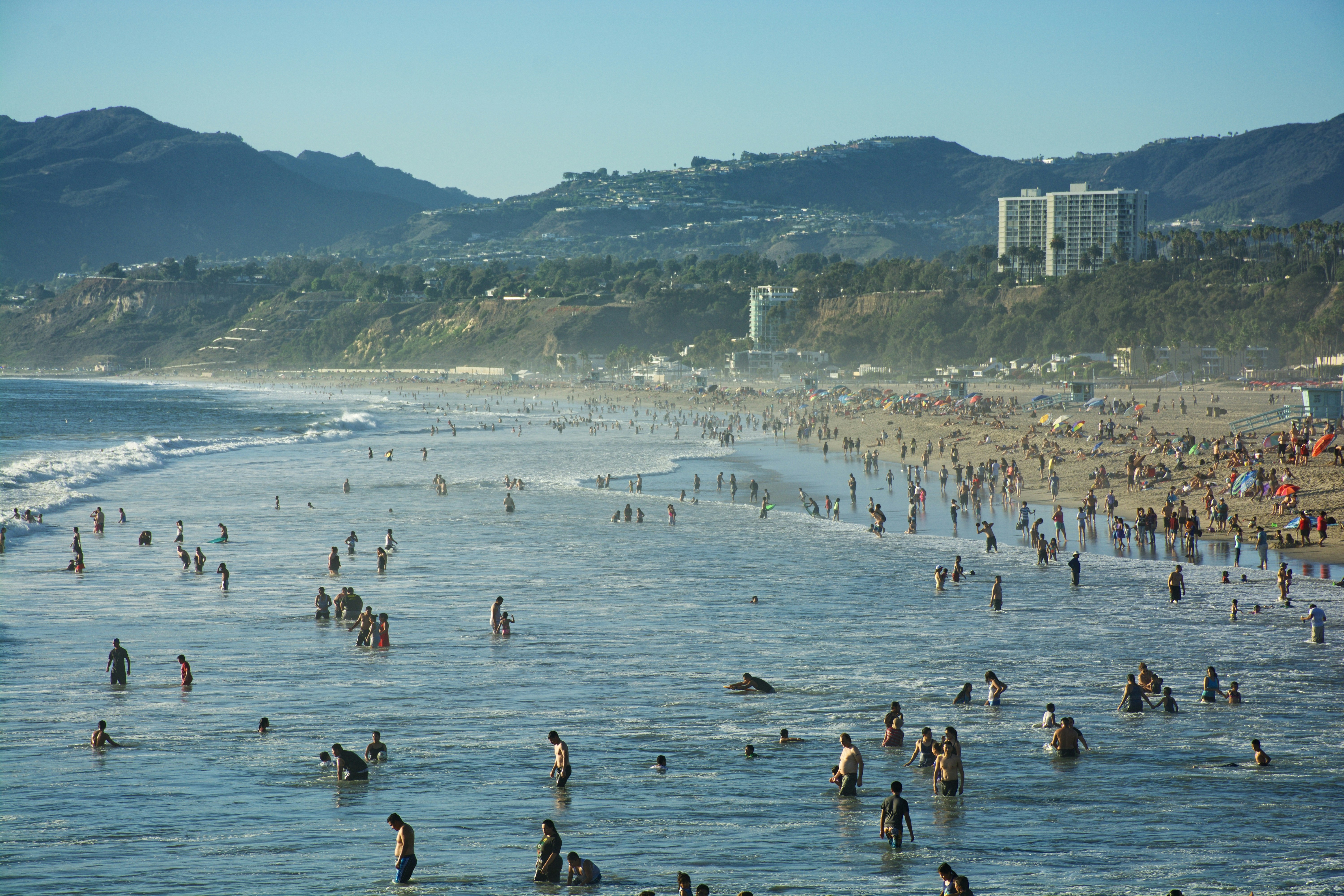 Vibrant beach scene filled with people enjoying the water and sun, framed by distant mountains and coastal buildings.