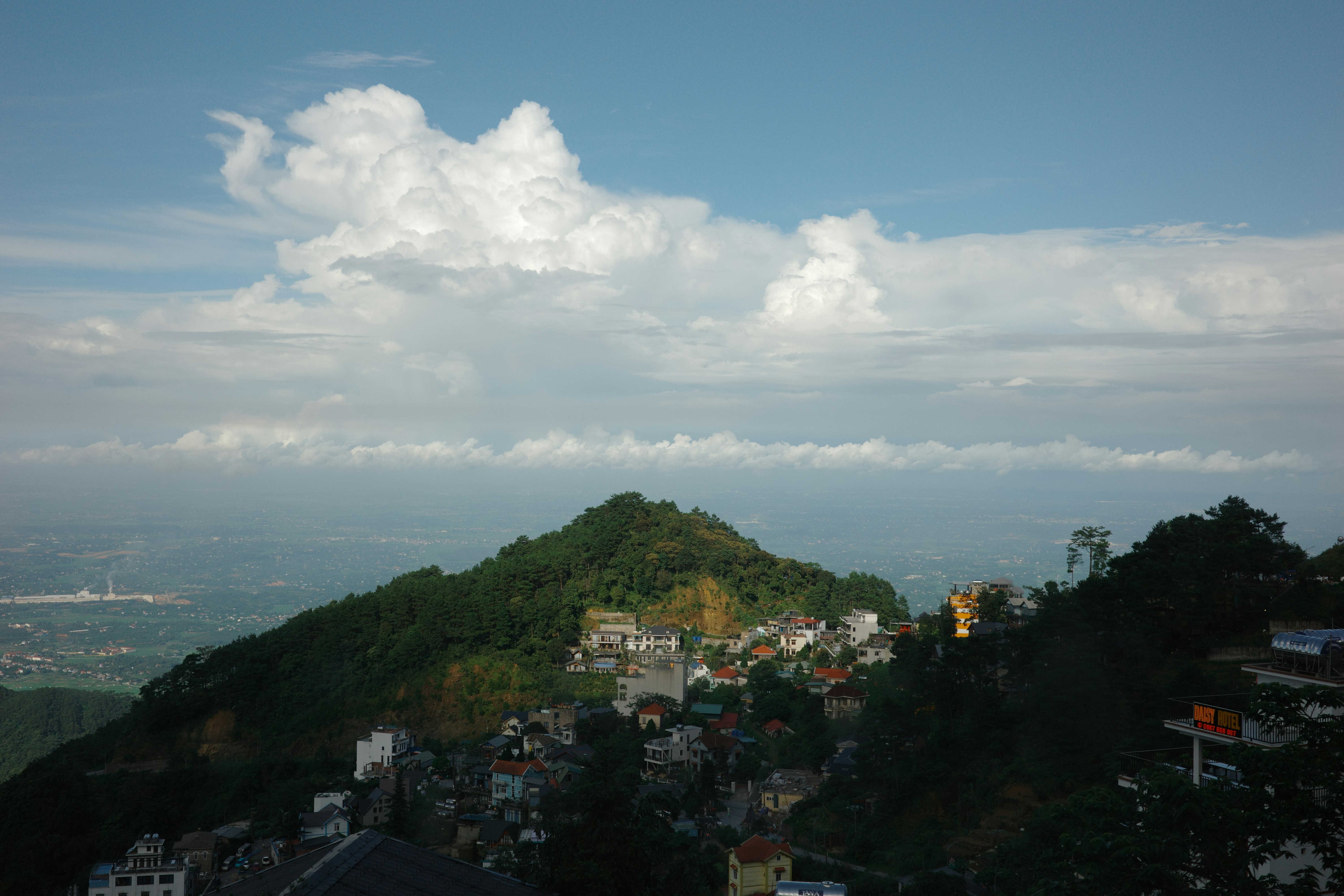 Lush green hills rise above a quaint village, framed by expansive clouds in a tranquil sky. The scene captures a serene mountainous landscape.