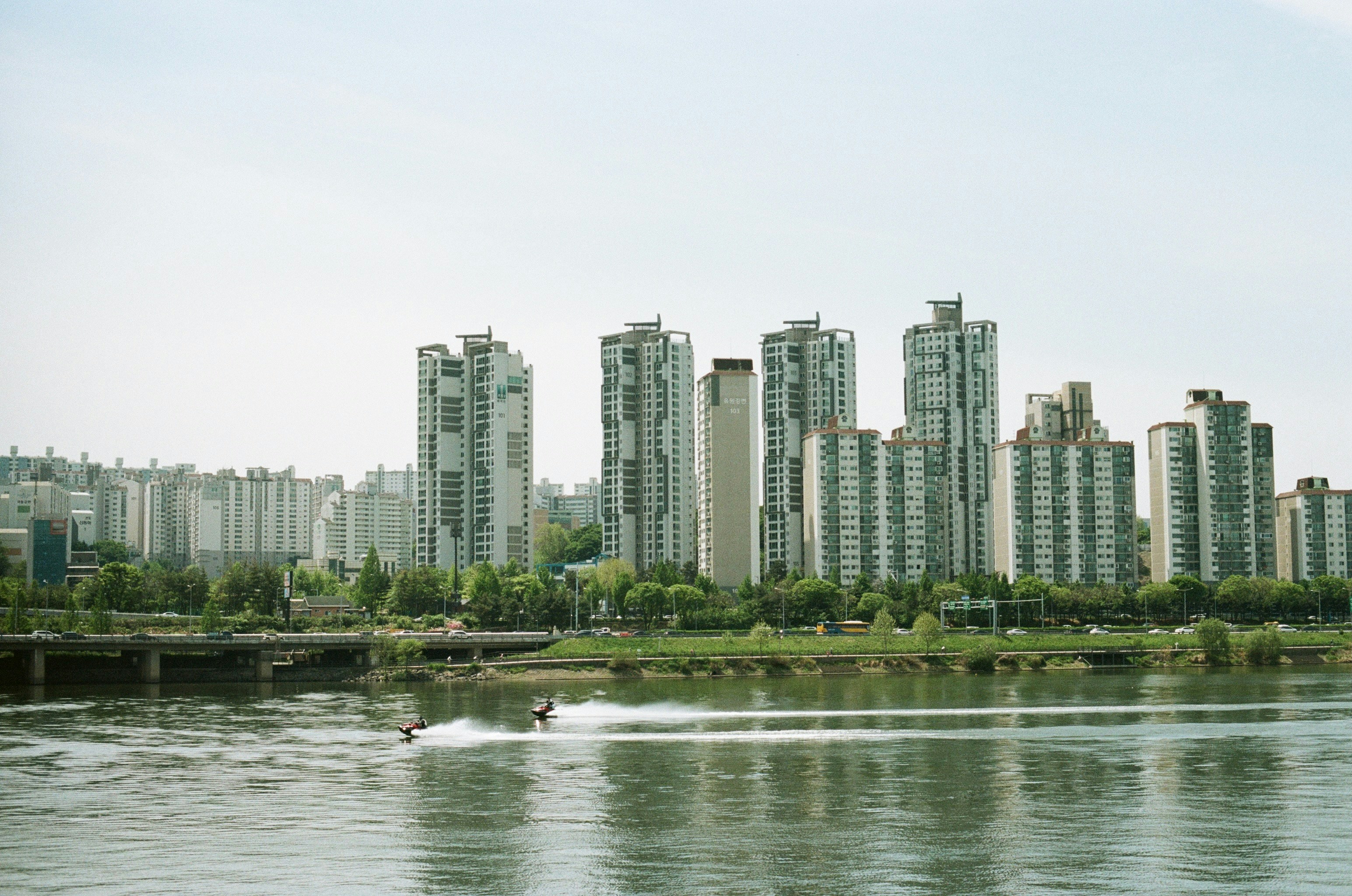 City skyline along a calm river with two water-skiers creating white wakes; high-rise residential towers line the embankment.