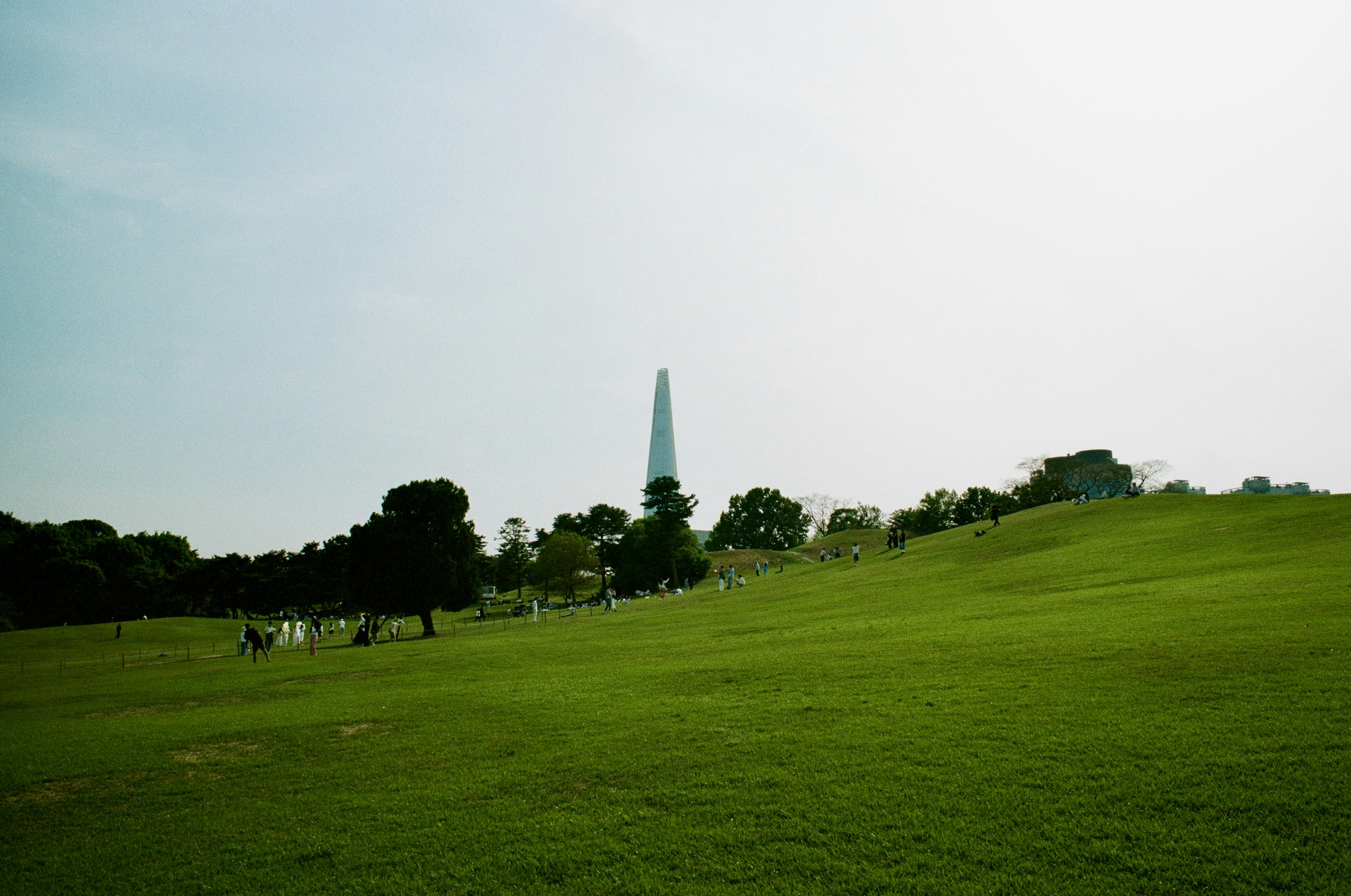 Grassy park slope with a distant white obelisk and small figures scattered across the hill under a clear blue sky.