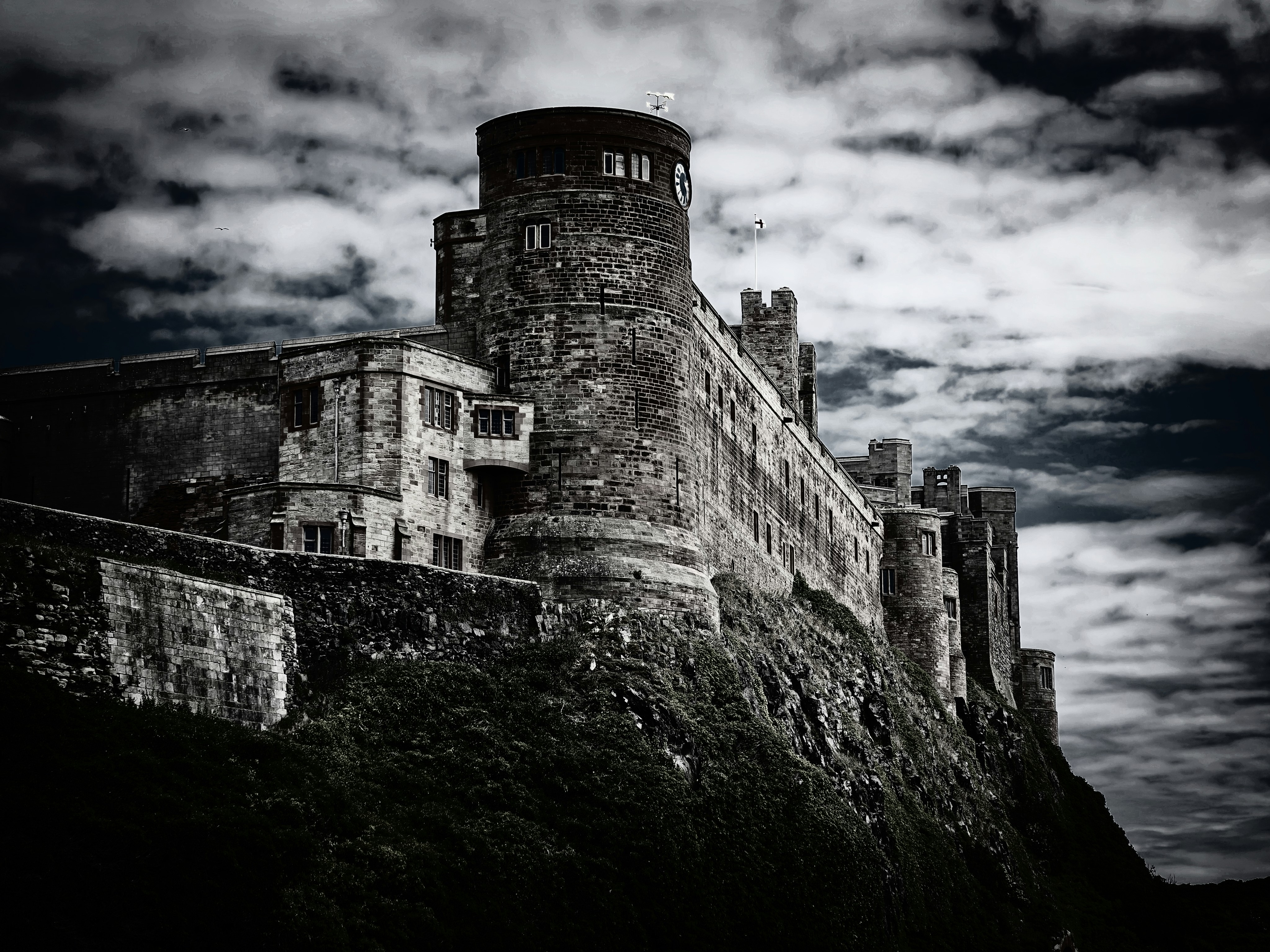 Black-and-white photograph of a medieval fortress perched on a rocky cliff under a turbulent, cloud-filled sky.