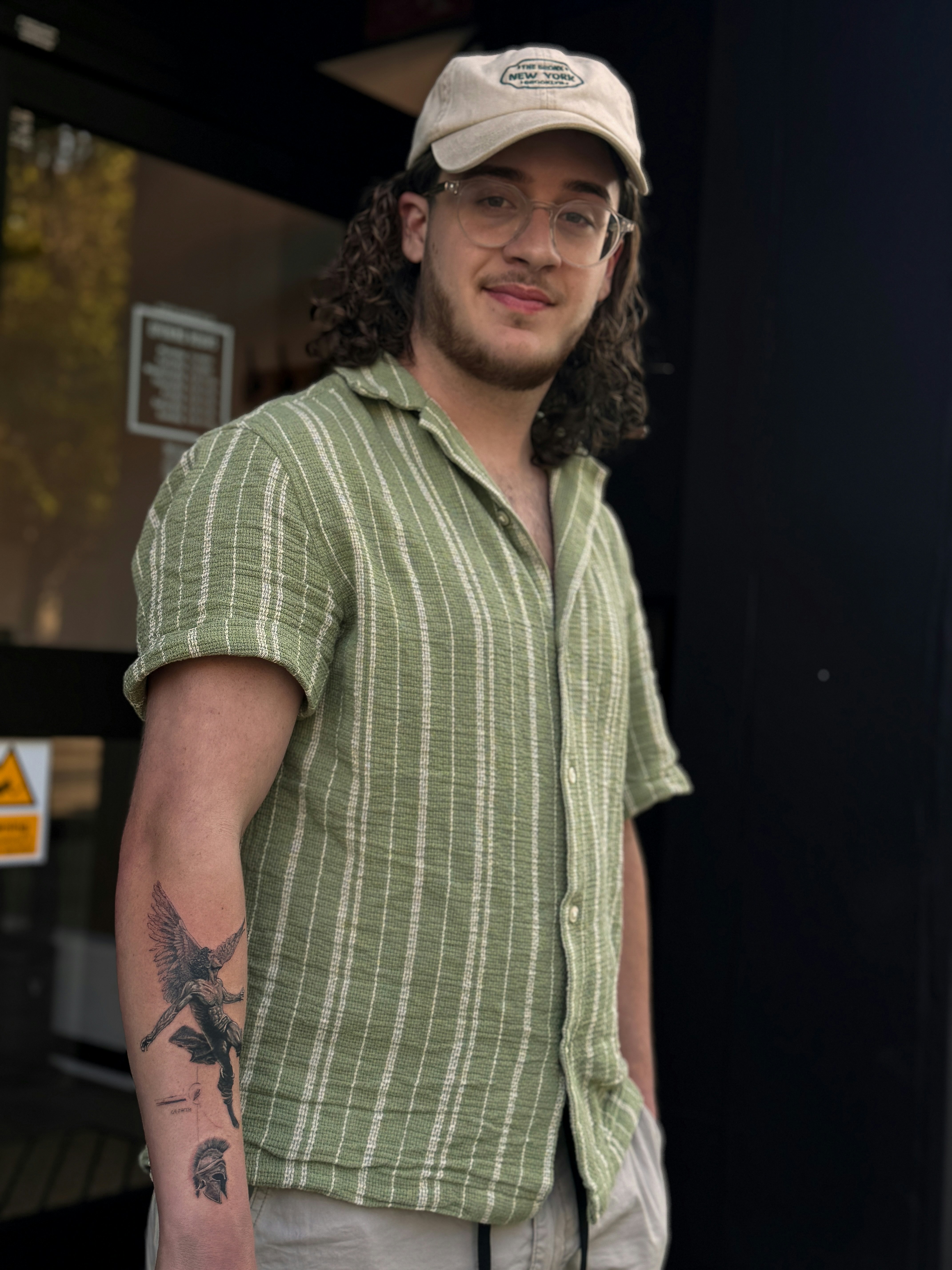 Young man in a green striped shirt and cap stands confidently outside a building, showcasing a detailed tattoo on his arm.