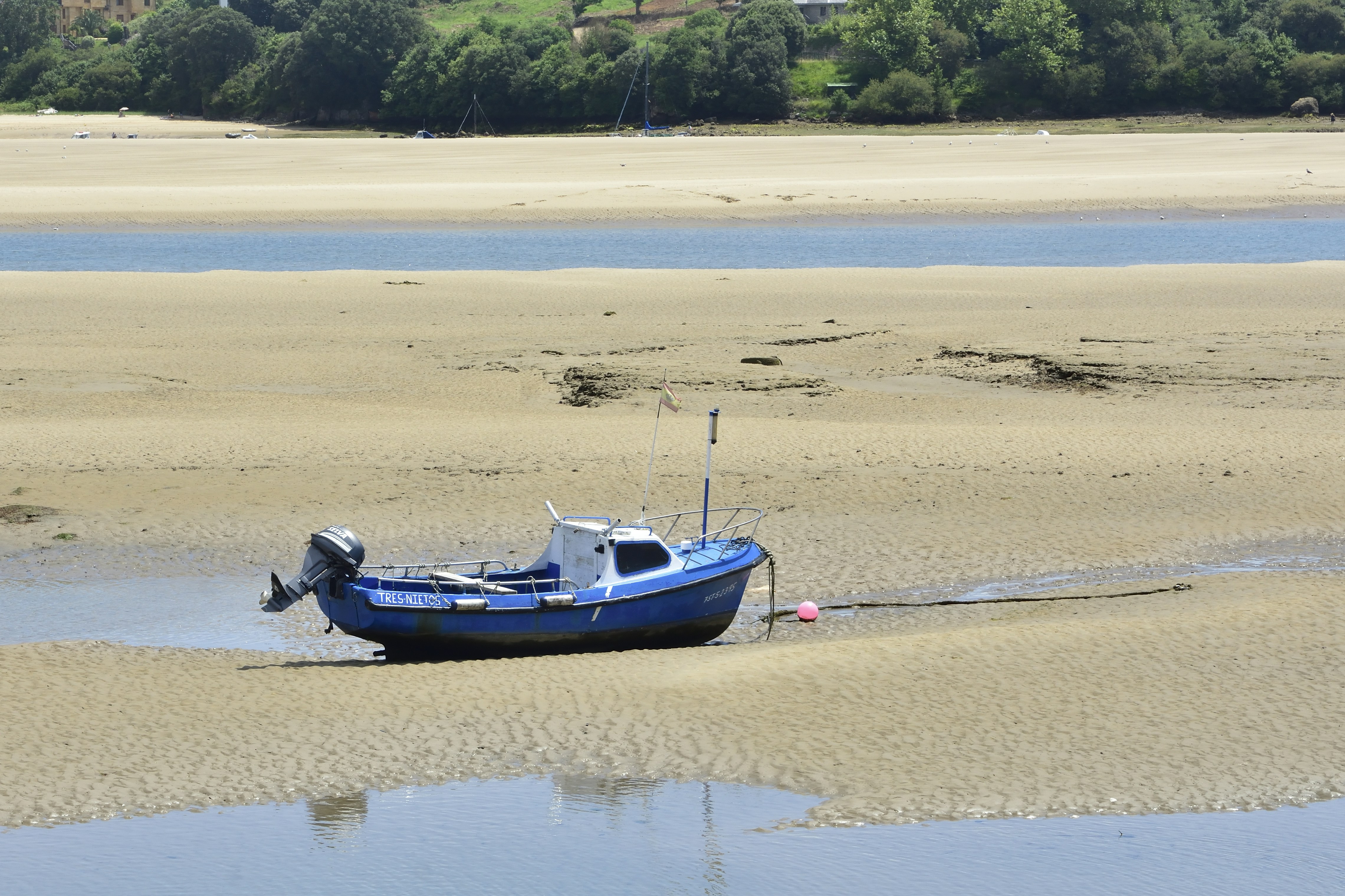 Blue boat rests on sandy shore with shallow water and lush greenery in the background.