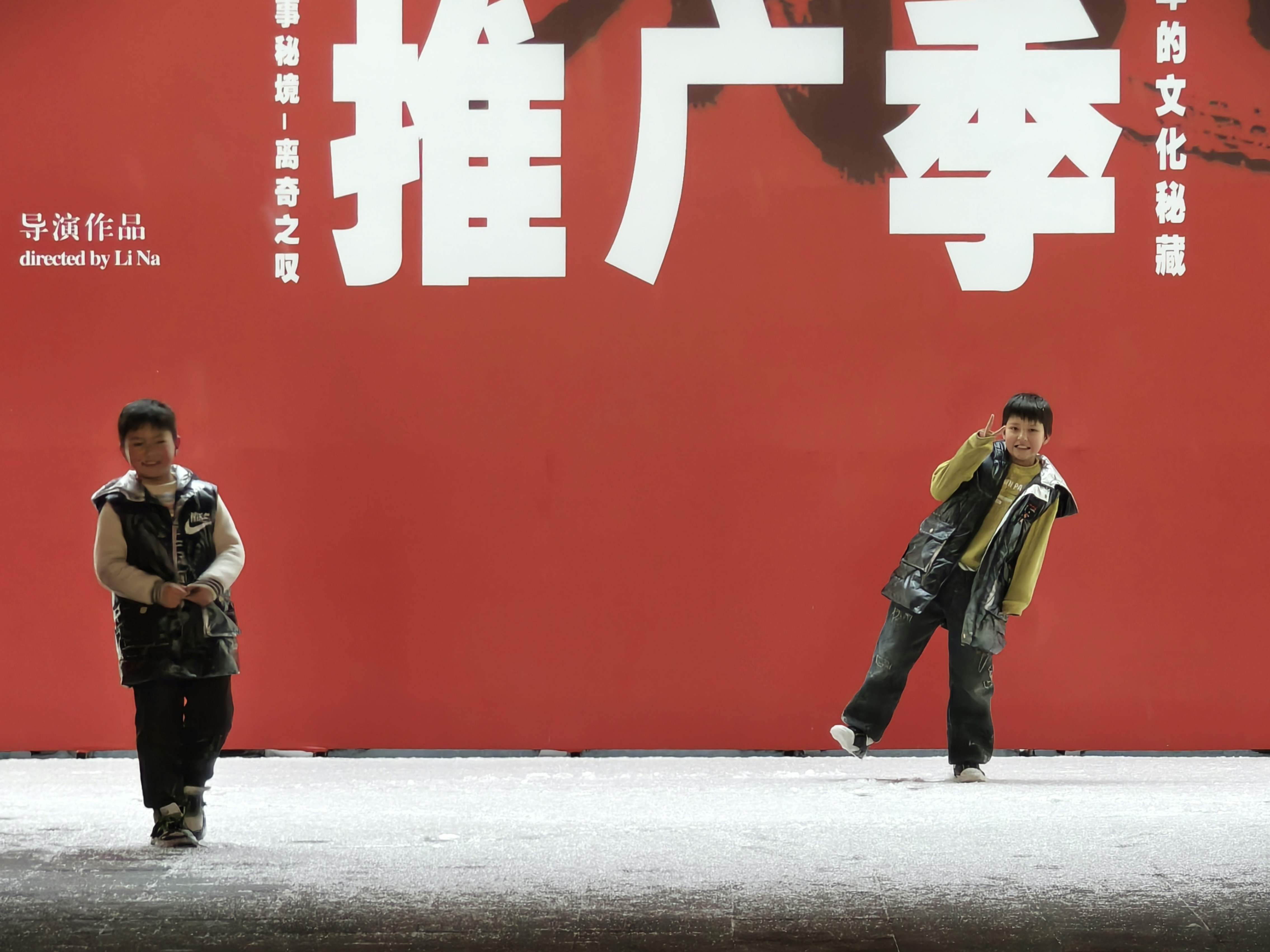 Two children stand on a snowy surface in front of a bold red wall with large white Chinese characters, captured in a candid street photograph.