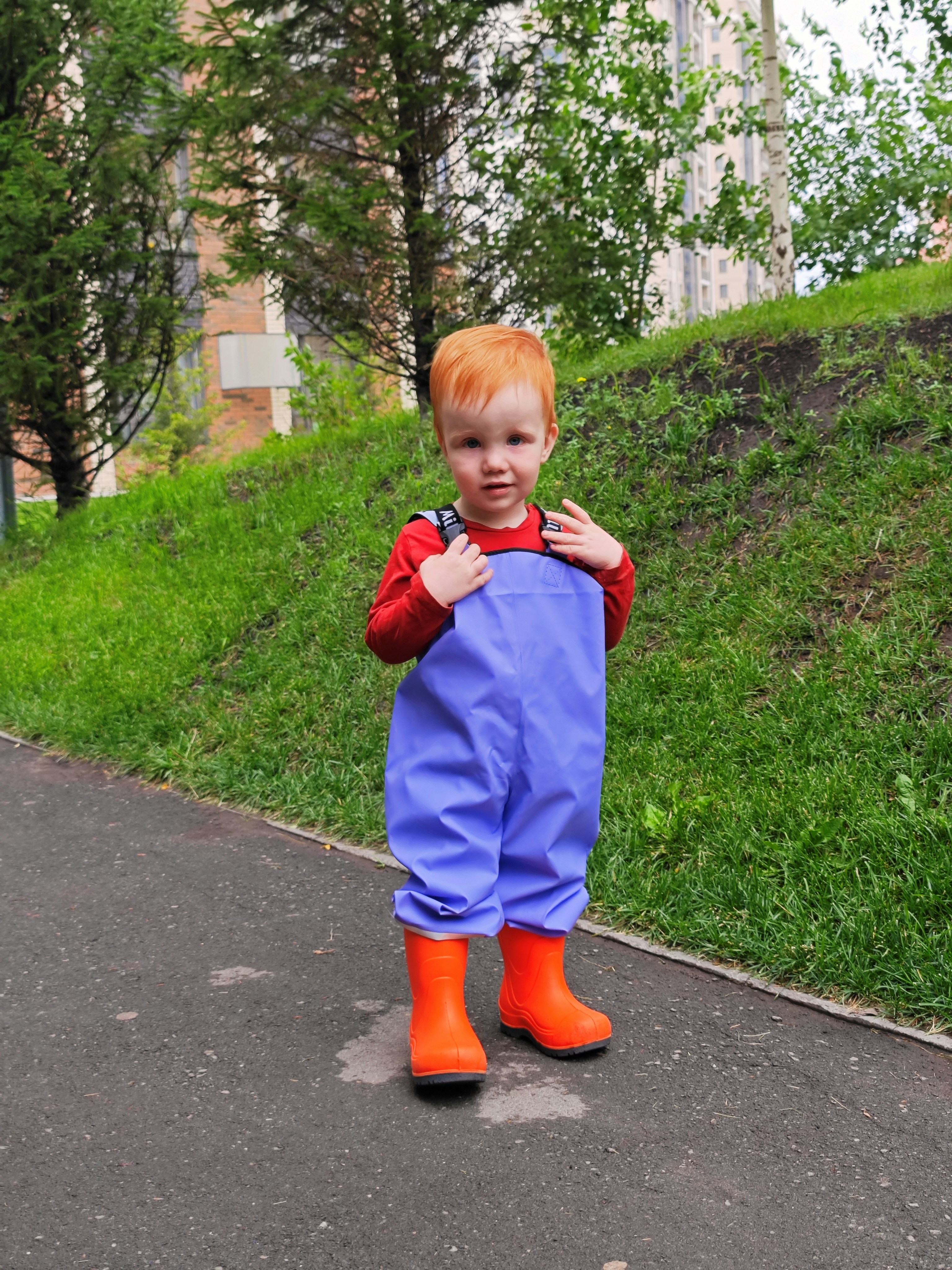A toddler with bright orange hair stands on a park path wearing a red long-sleeve shirt, blue overalls, and vivid orange boots, with a grassy slope and trees in the background. This photograph captures a candid urban moment.