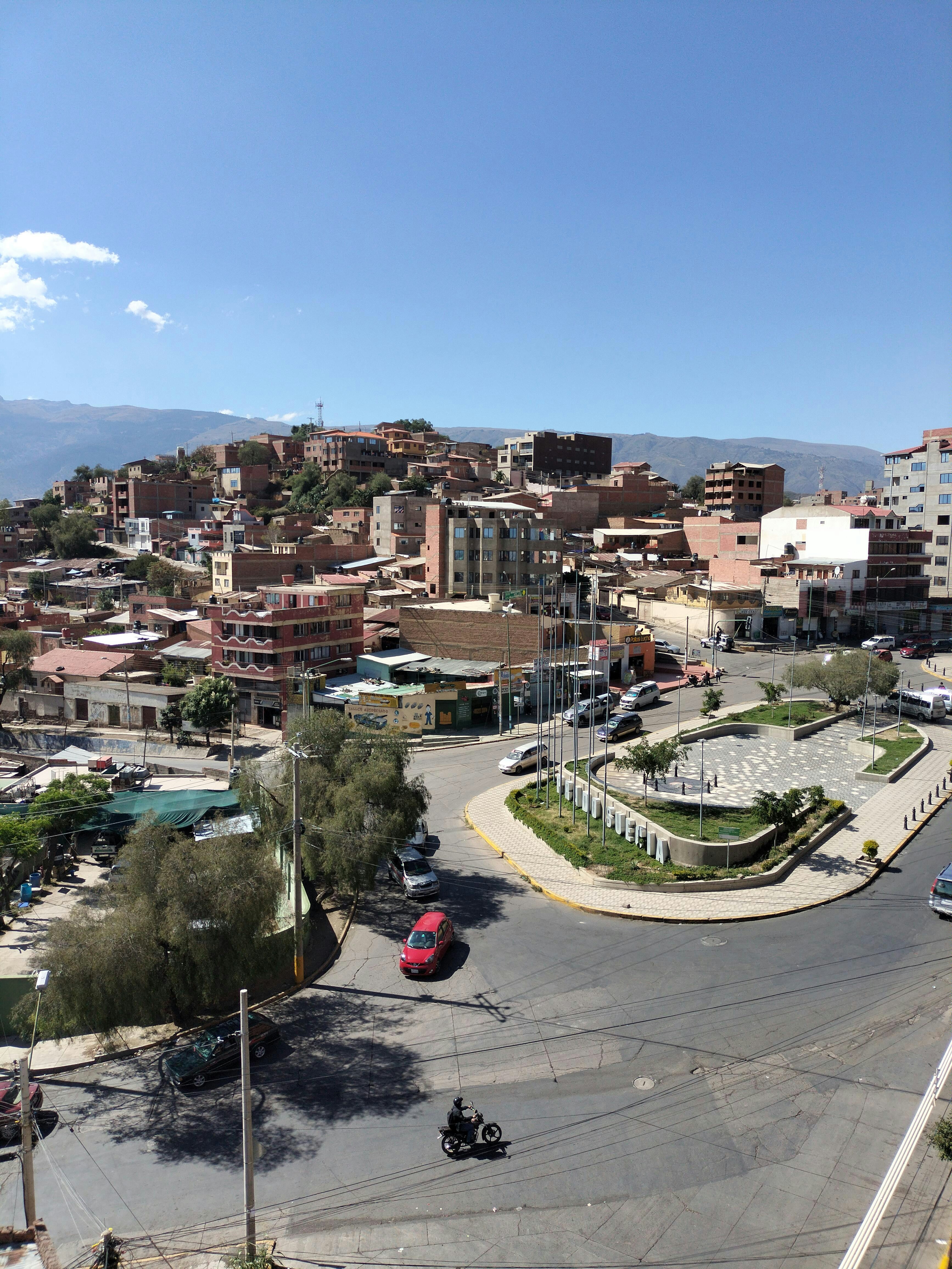 Aerial cityscape photograph from a high vantage, featuring a circular roundabout, dense brick buildings, and distant hills beneath a clear blue sky.