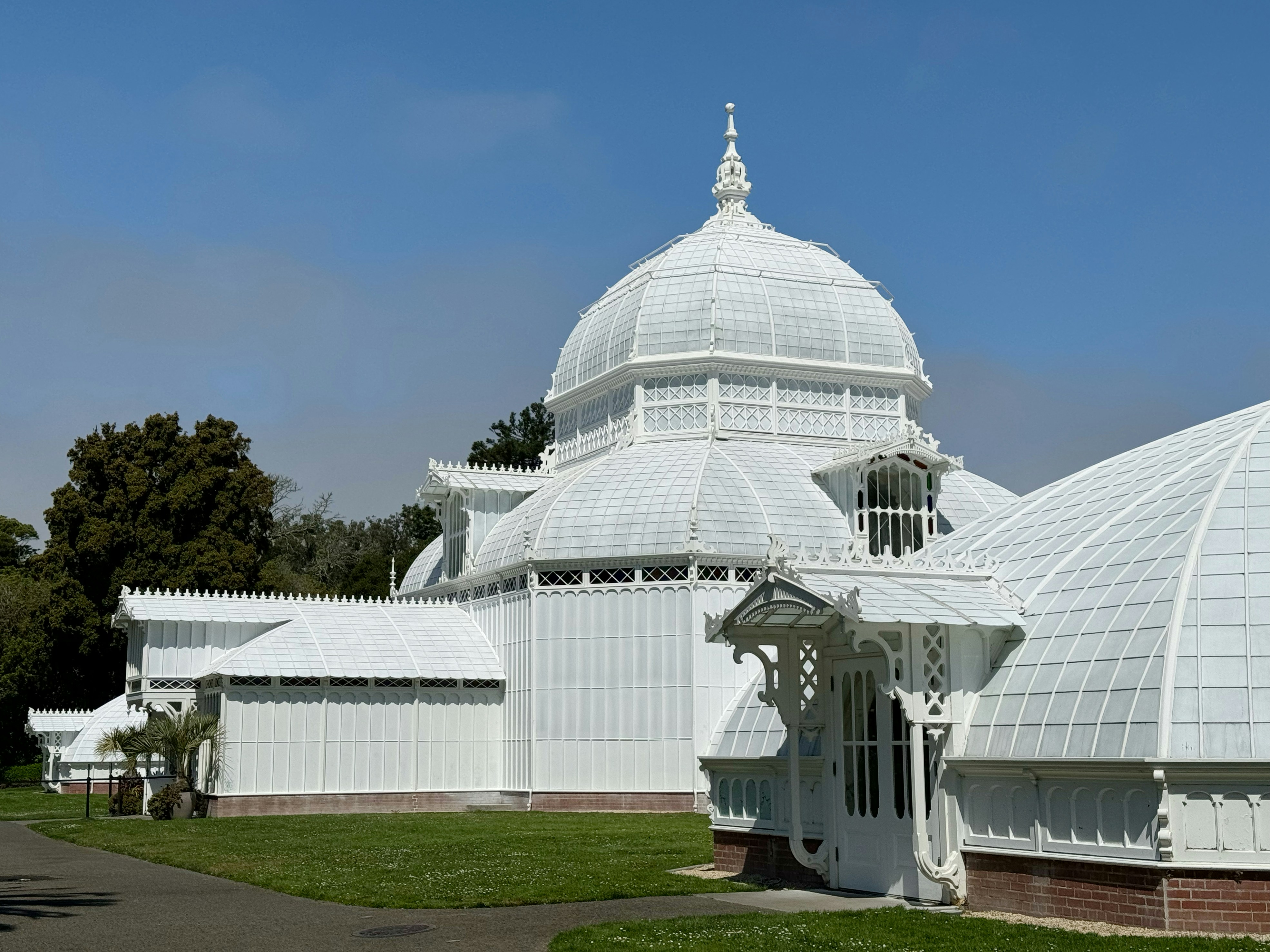 A Victorian-style conservatory featuring intricate glass architecture, surrounded by lush greenery in a serene park setting.