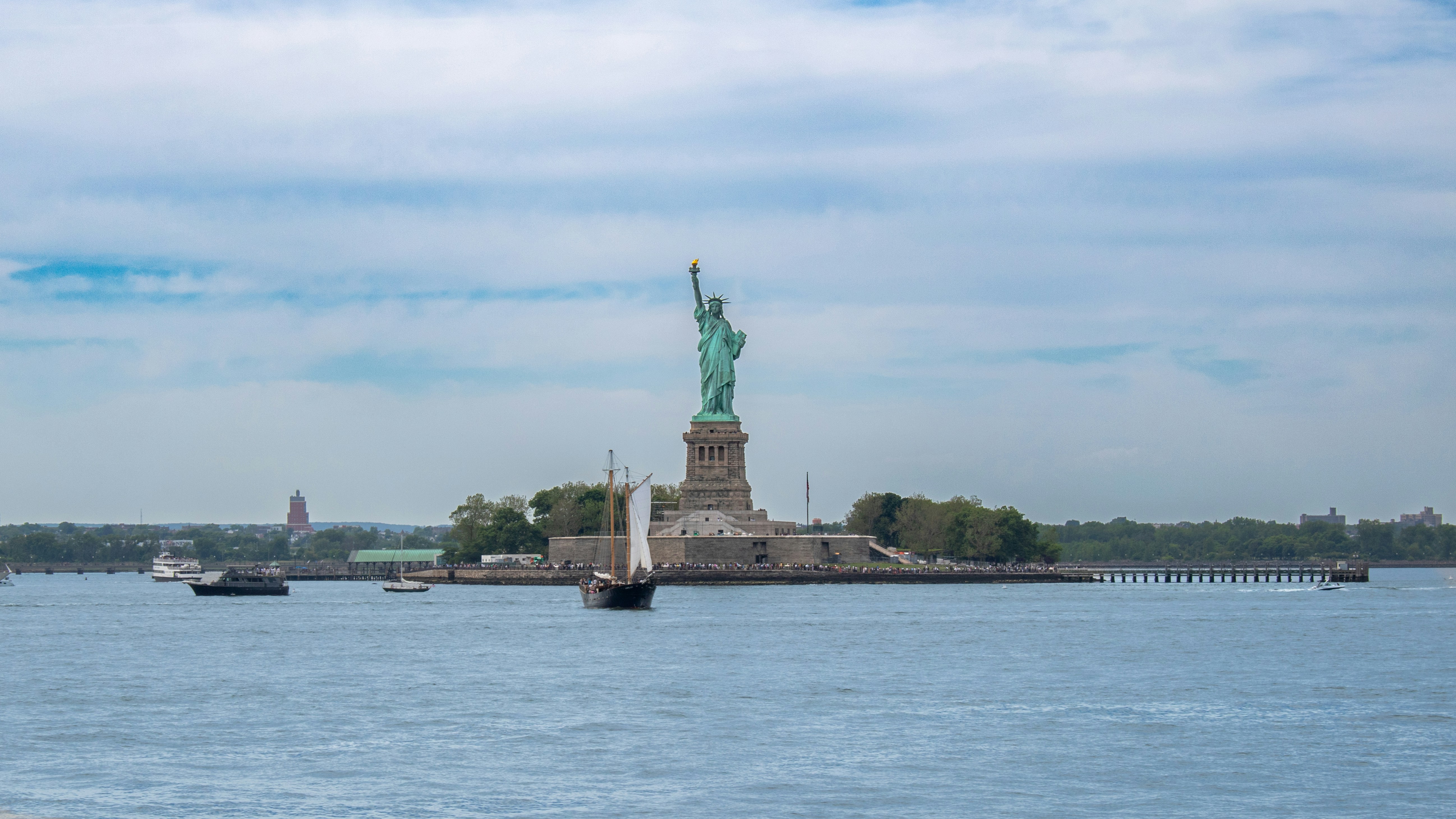 The Statue of Liberty stands tall with sailboats passing in the foreground on a calm day.