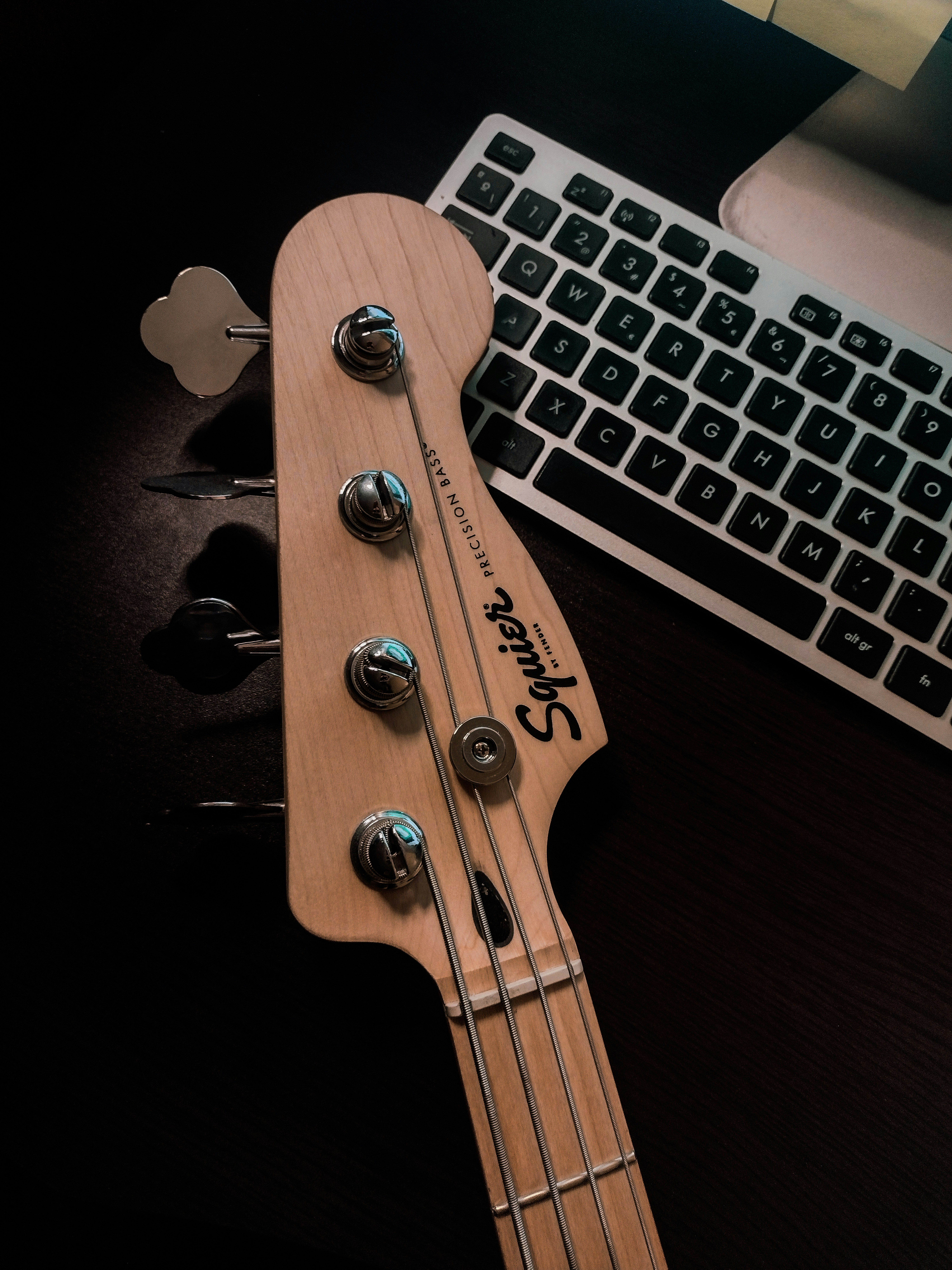 Close-up photograph of a Squier guitar headstock with tuning pegs and logo resting on a dark desk beside a laptop. The shallow depth of field keeps the headstock in focus while the keyboard fades softly.