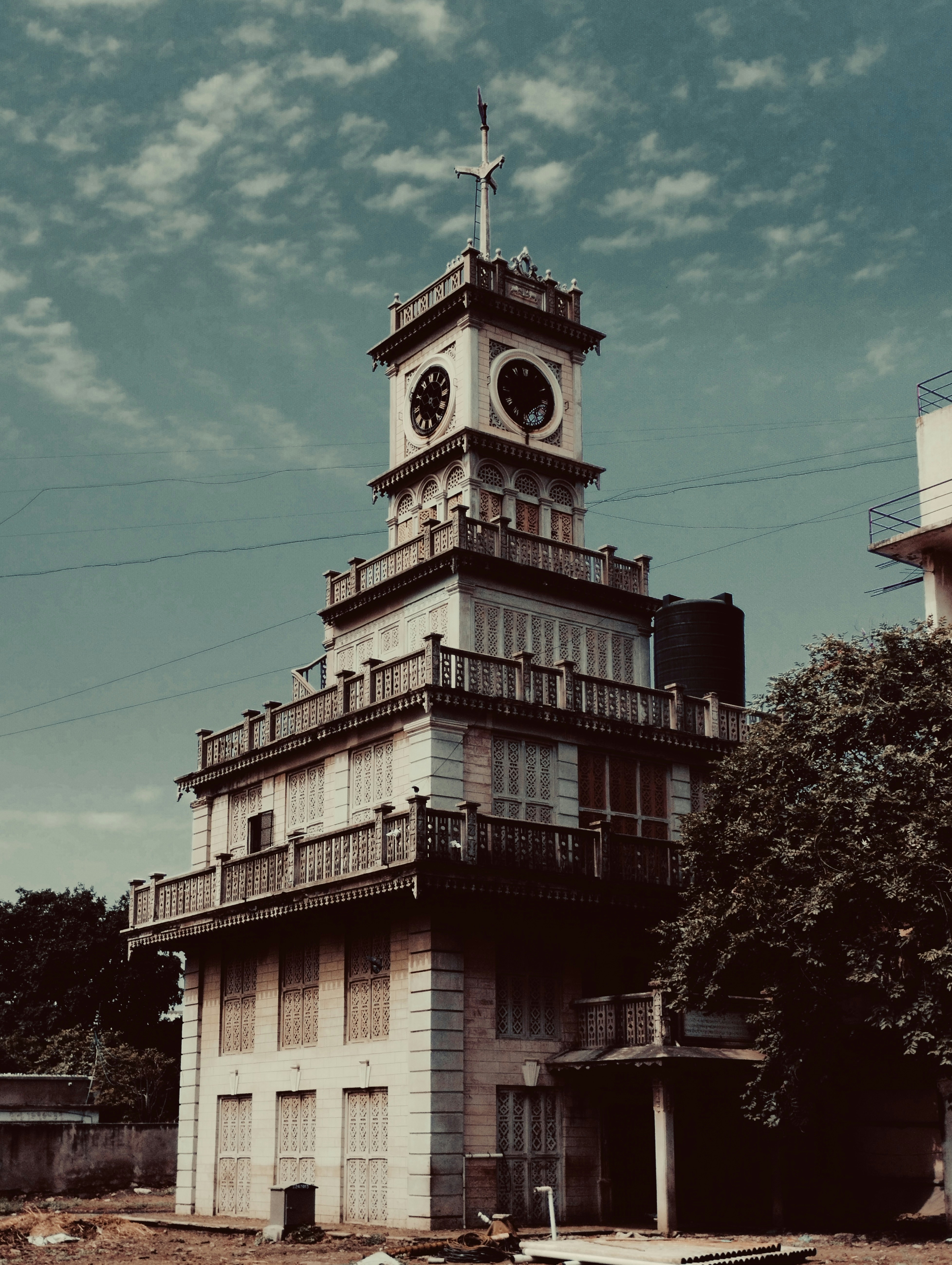 A multi-tiered clocktower rises above a quiet street, framed by a teal sky and surrounding trees.