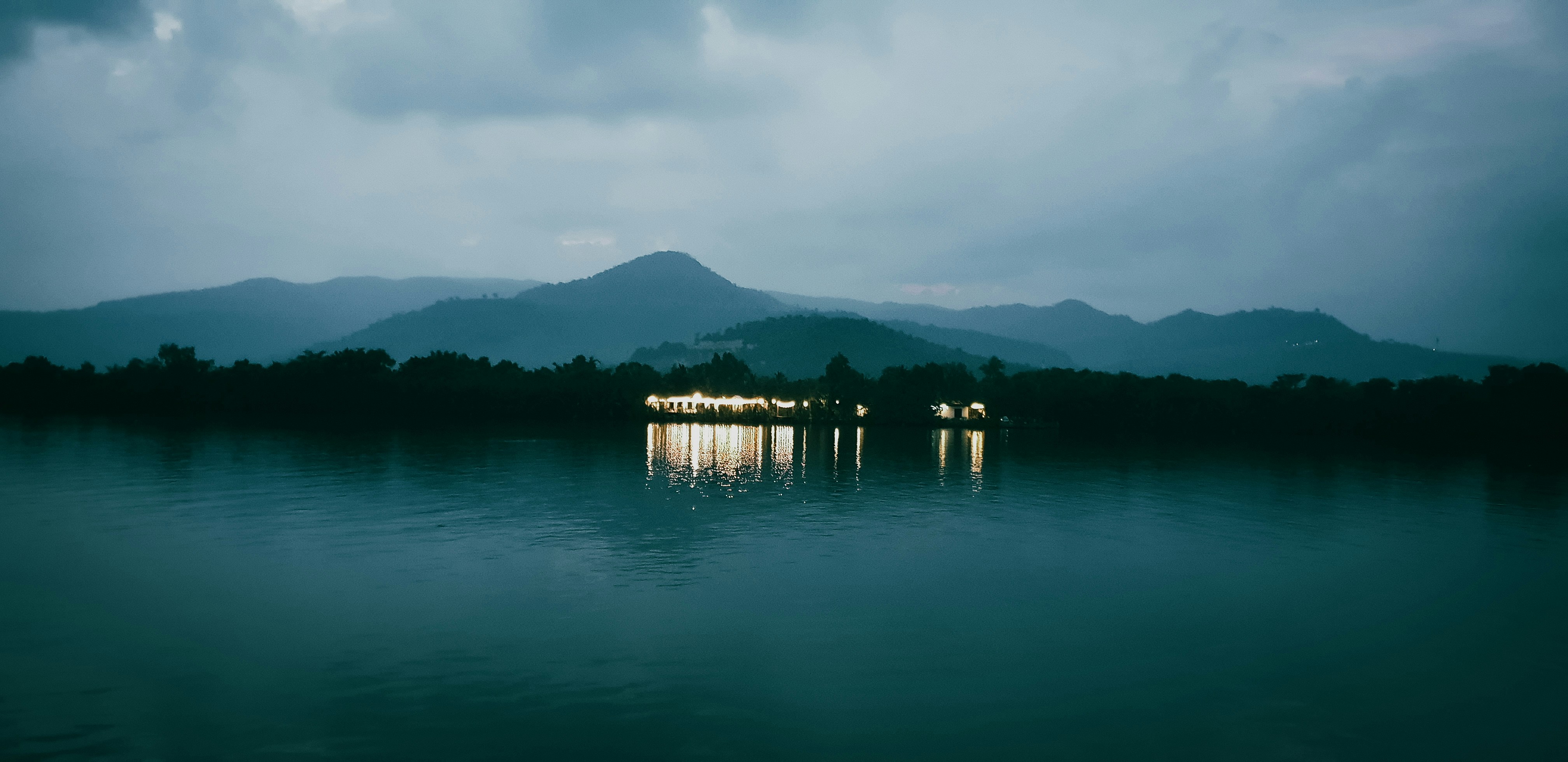Mountains silhouetted against a cloudy sky with lights reflecting on a tranquil lake.