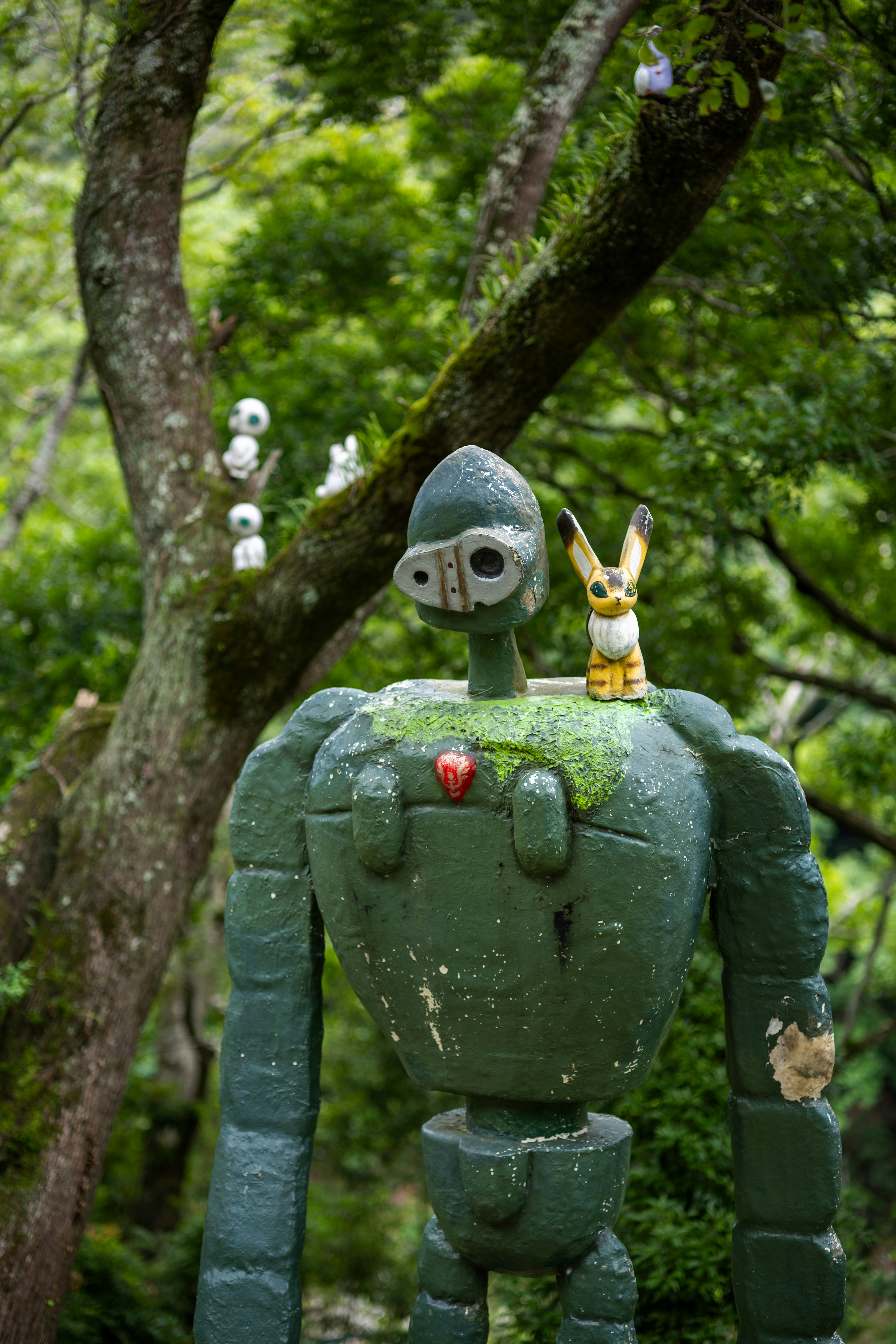 A moss-covered green robot sculpture dominates a forest clearing, with a small yellow rabbit figure perched on its shoulder. In the background, a tree bears a row of small white globe figures.