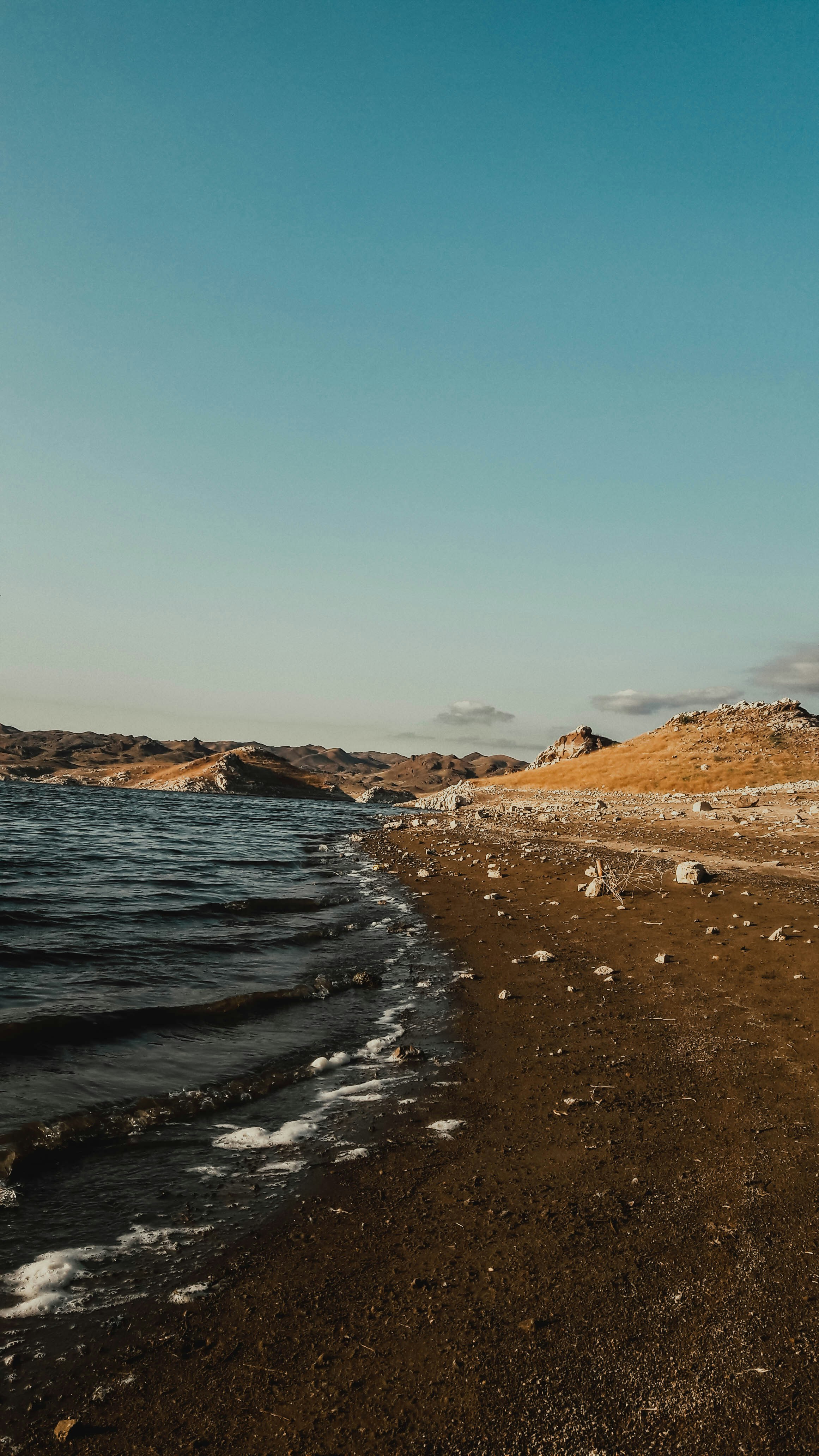 Desolate coastline with dark water washing a pebble beach and rust-colored hills in the distance beneath a pale gradient sky.