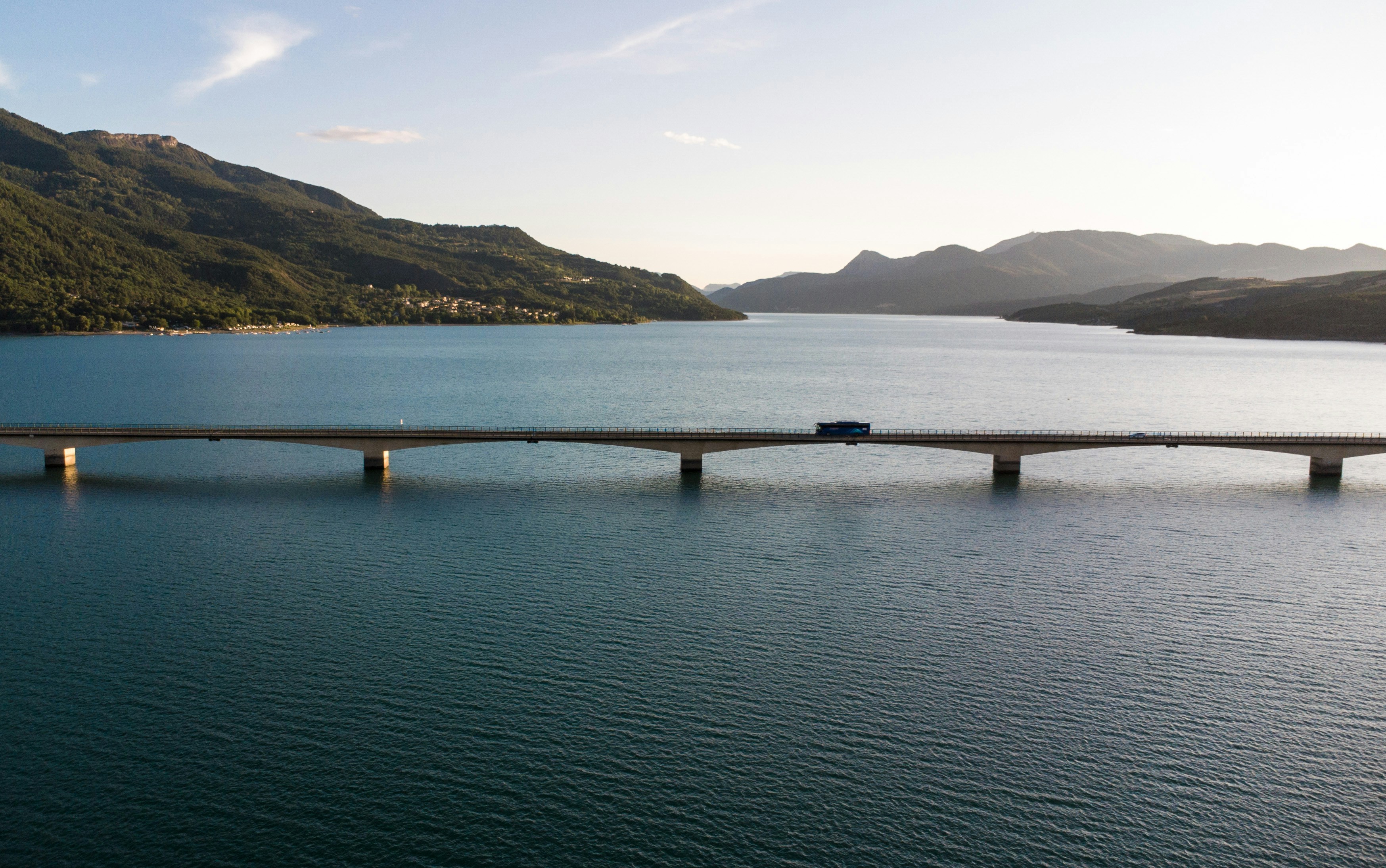 Long bridge spans a tranquil fjord with mountains in the distance, bathed in soft golden light. A lone car crosses the bridge, highlighting scale and quiet movement.