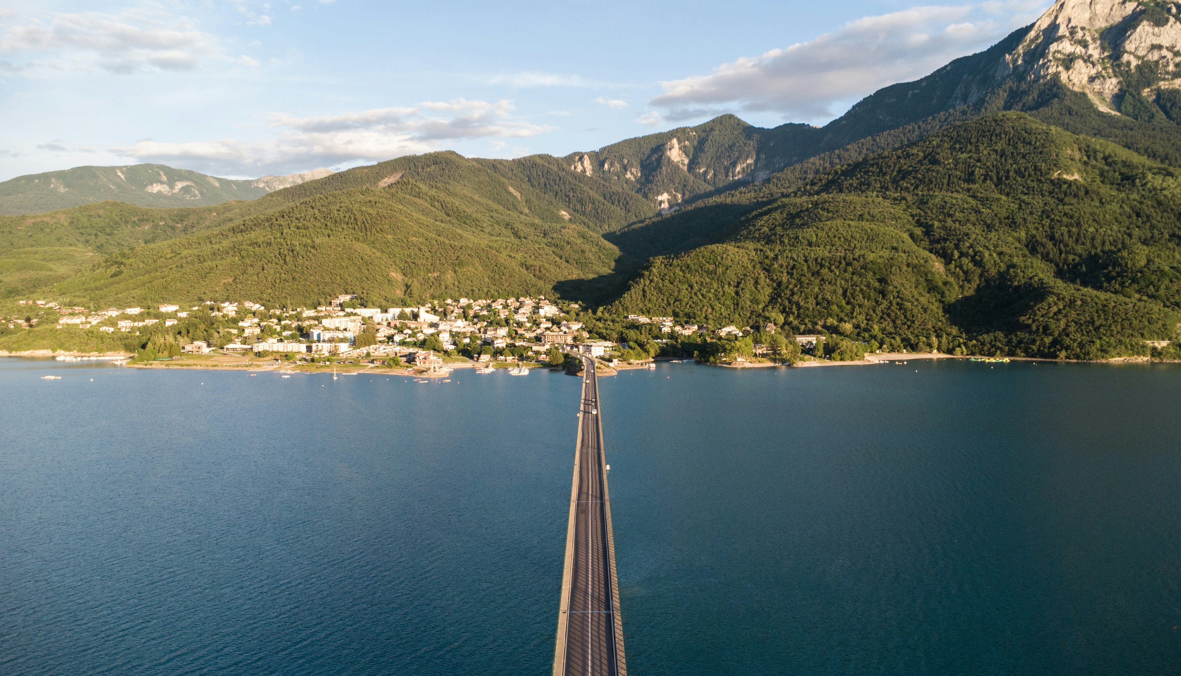 Drone photograph of a long bridge extending to a coastal village framed by forested mountains.
