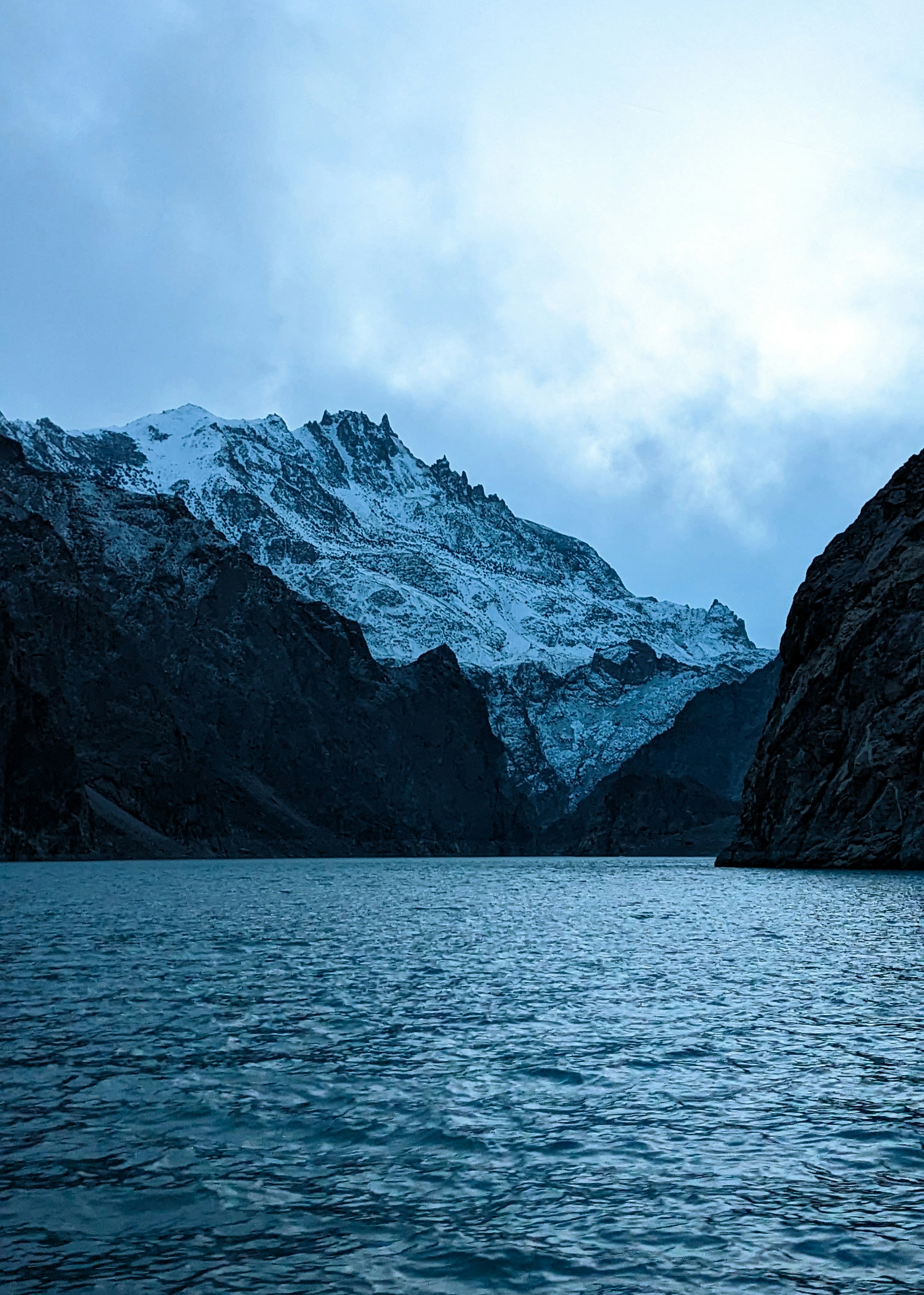 Snow-capped mountains loom over a tranquil lake, shrouded in a soft blue haze. The rugged terrain reflects the calm waters below.