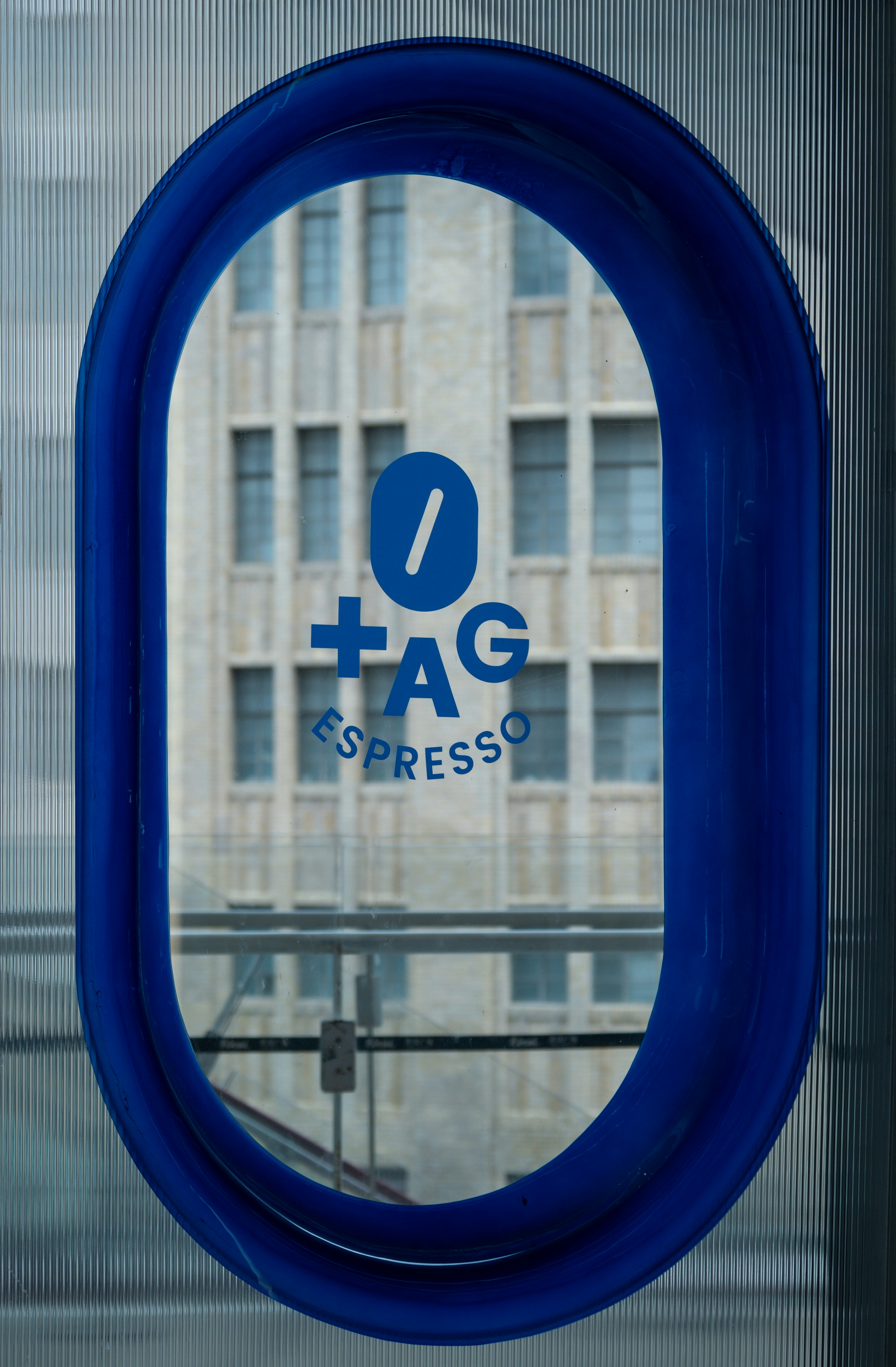 Blue oval frame surrounds a bold espresso logo on glass, with a distant brick building visible through the pane.
