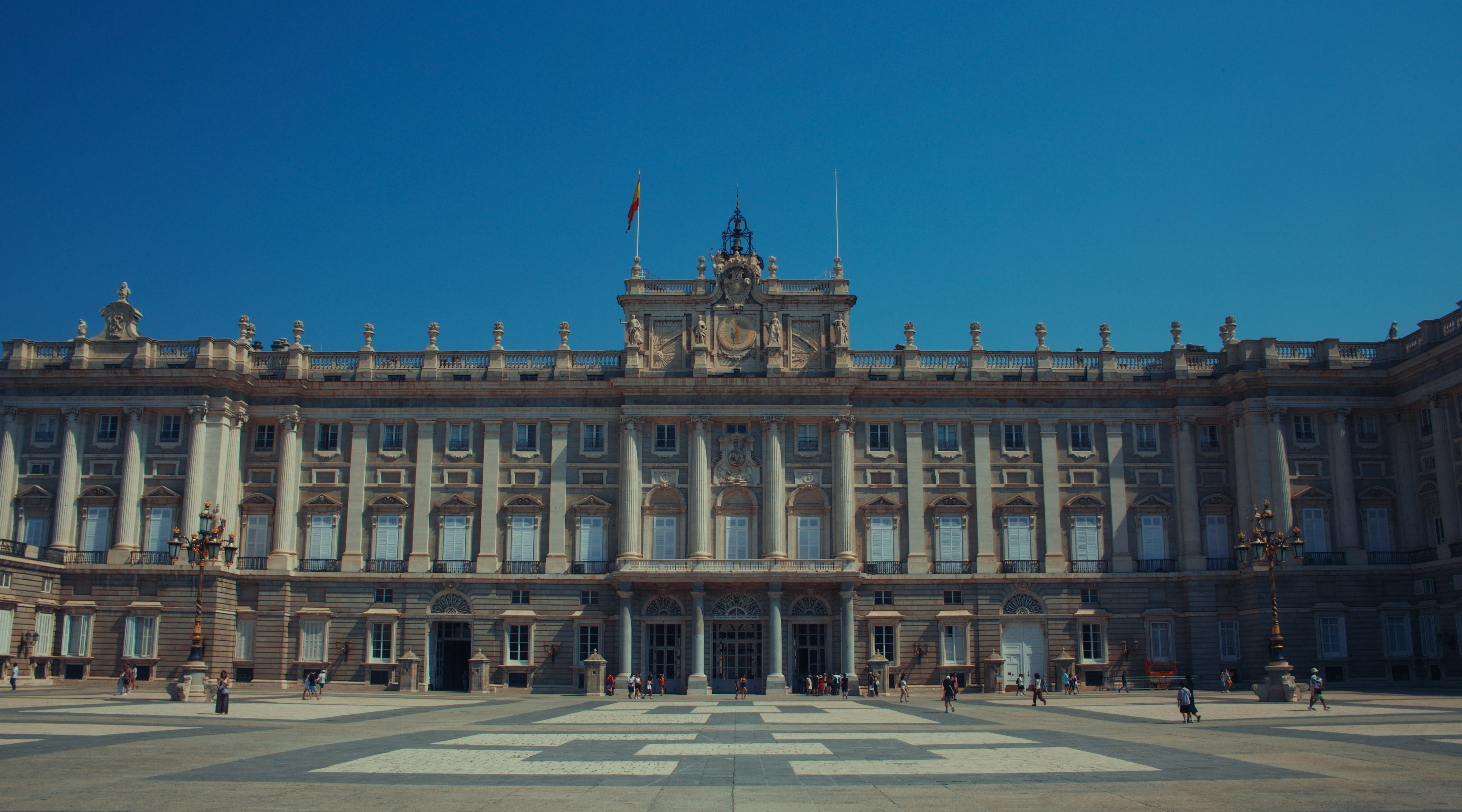 The grand facade of a historic palace showcases intricate architectural details under a clear blue sky. Visitors stroll across the expansive plaza.