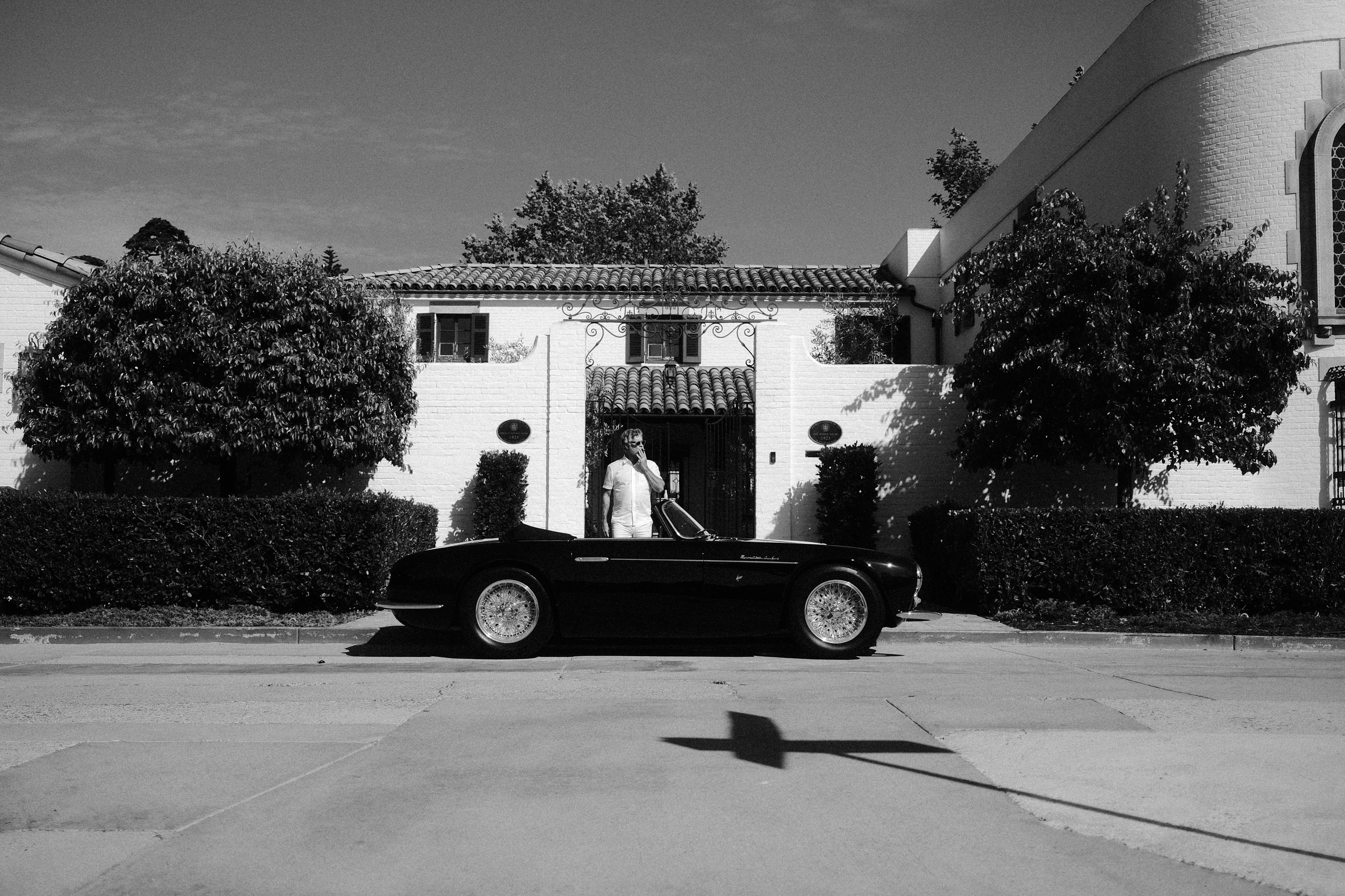 Man smoking cigar outside of home with a nice car in front of him.