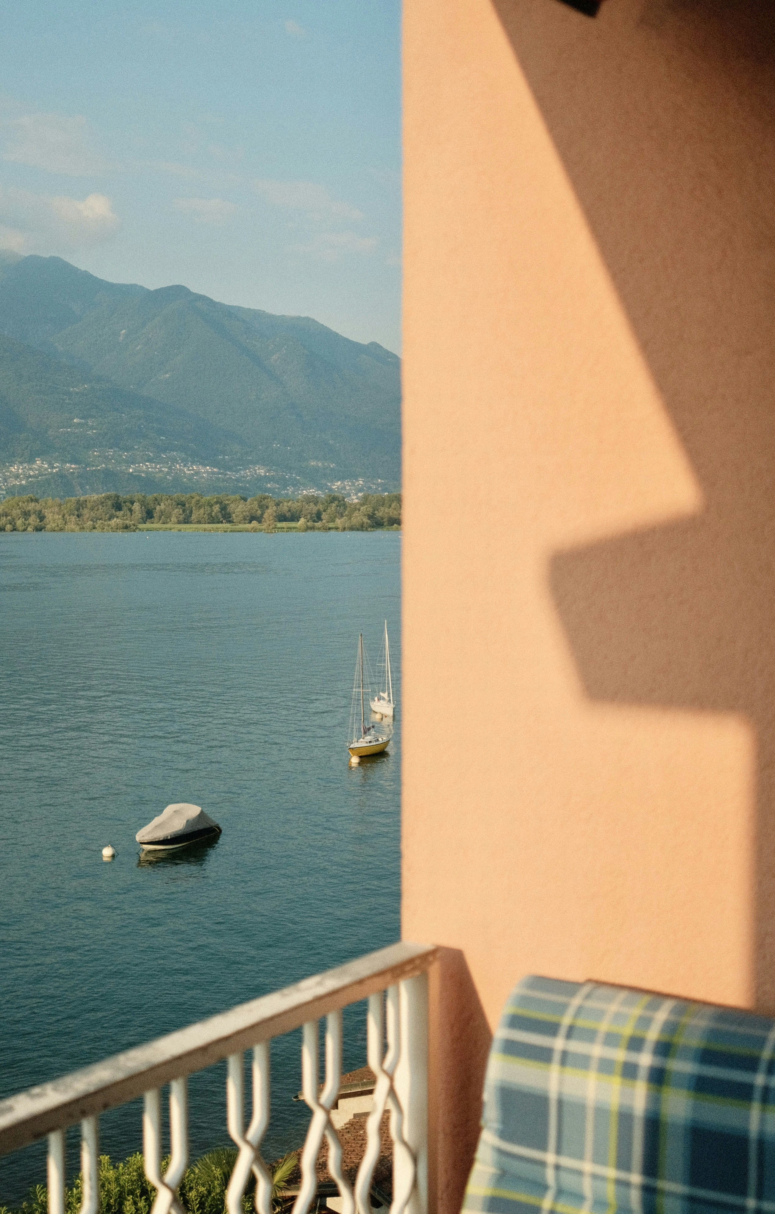 Enjoy a tranquil moment by Lago Maggiore: This image captures the serene beauty of the lake, framed by the warm tones of a Mediterranean structure. The play of light and shadow adds a touch of the Dolce Vita, highlighting the boats gently swaying in the water. The distant mountains provide a perfect backdrop, emphasizing the idyllic and picturesque nature of this Mediterranean paradise. | A balcony with a chair and a view of a lake