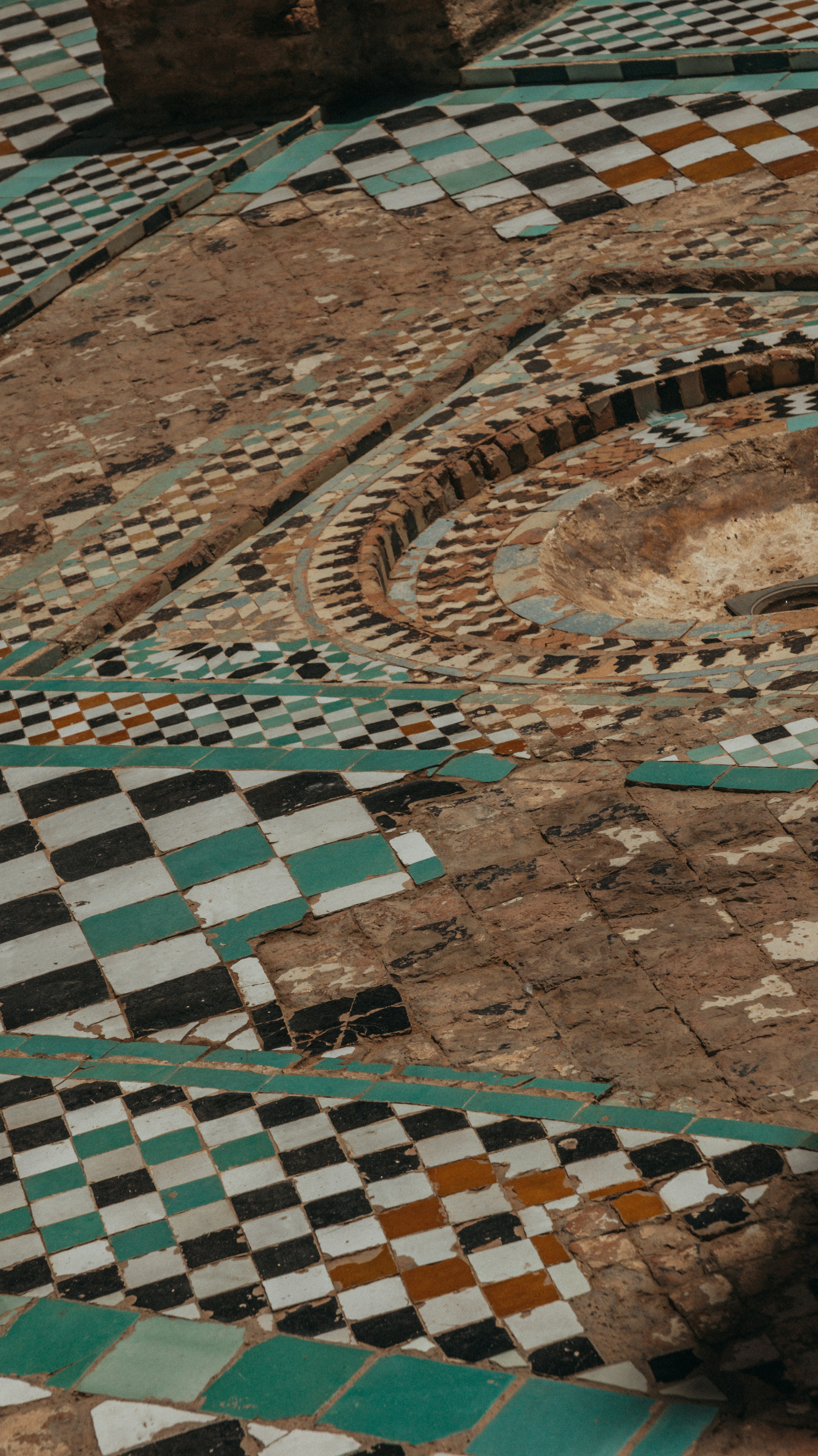 Close-up of a weathered mosaic tile floor with a circular brick feature and dirt, captured in warm, earthy tones.