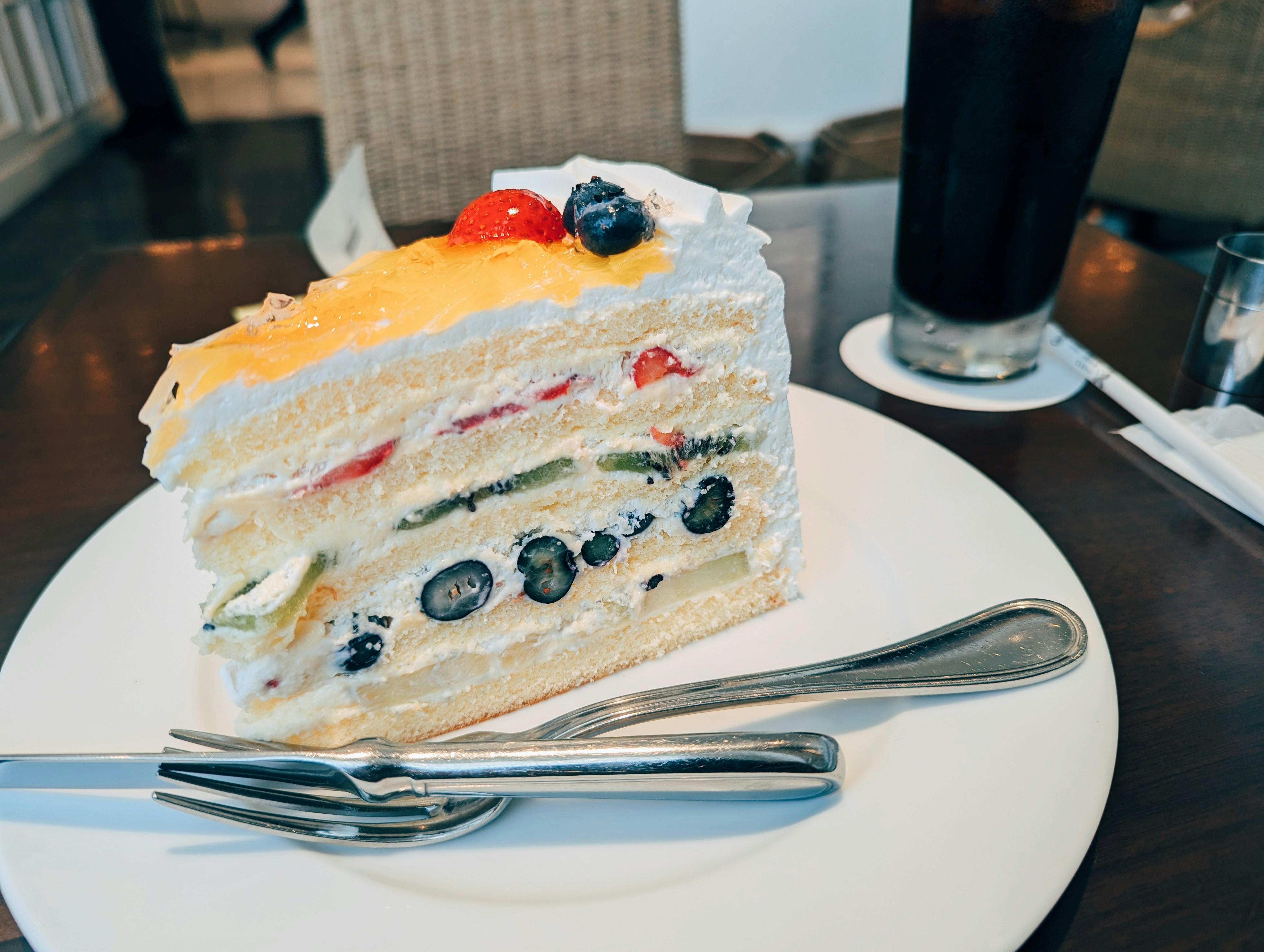 A slice of layered fruit cake with cream, blueberries, and strawberries rests on a white plate in a cozy café setting. A dark beverage and utensils complete the scene.
