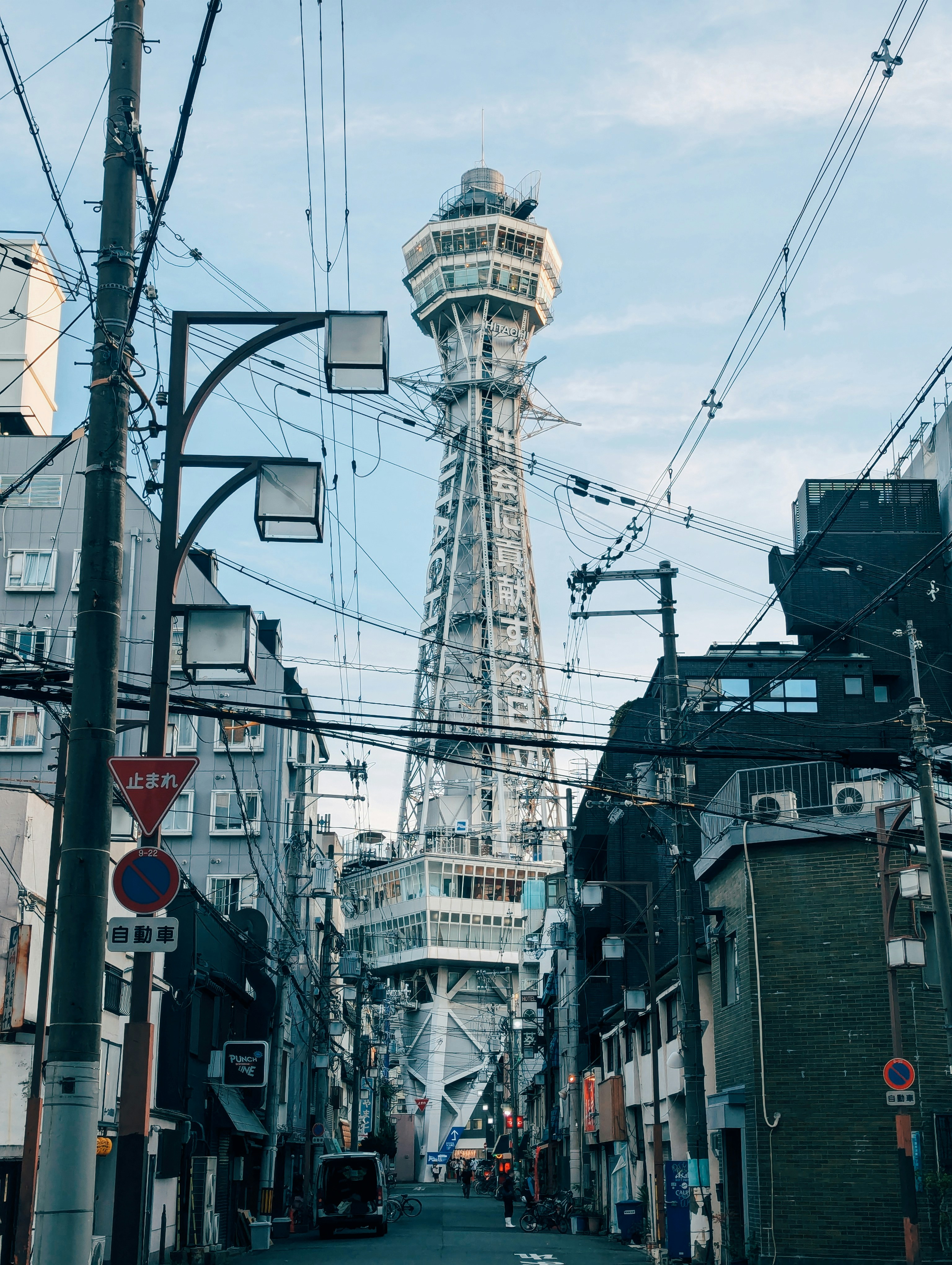 A narrow urban street framed by buildings and overhead cables leads toward a tall lattice observation tower in the distance.