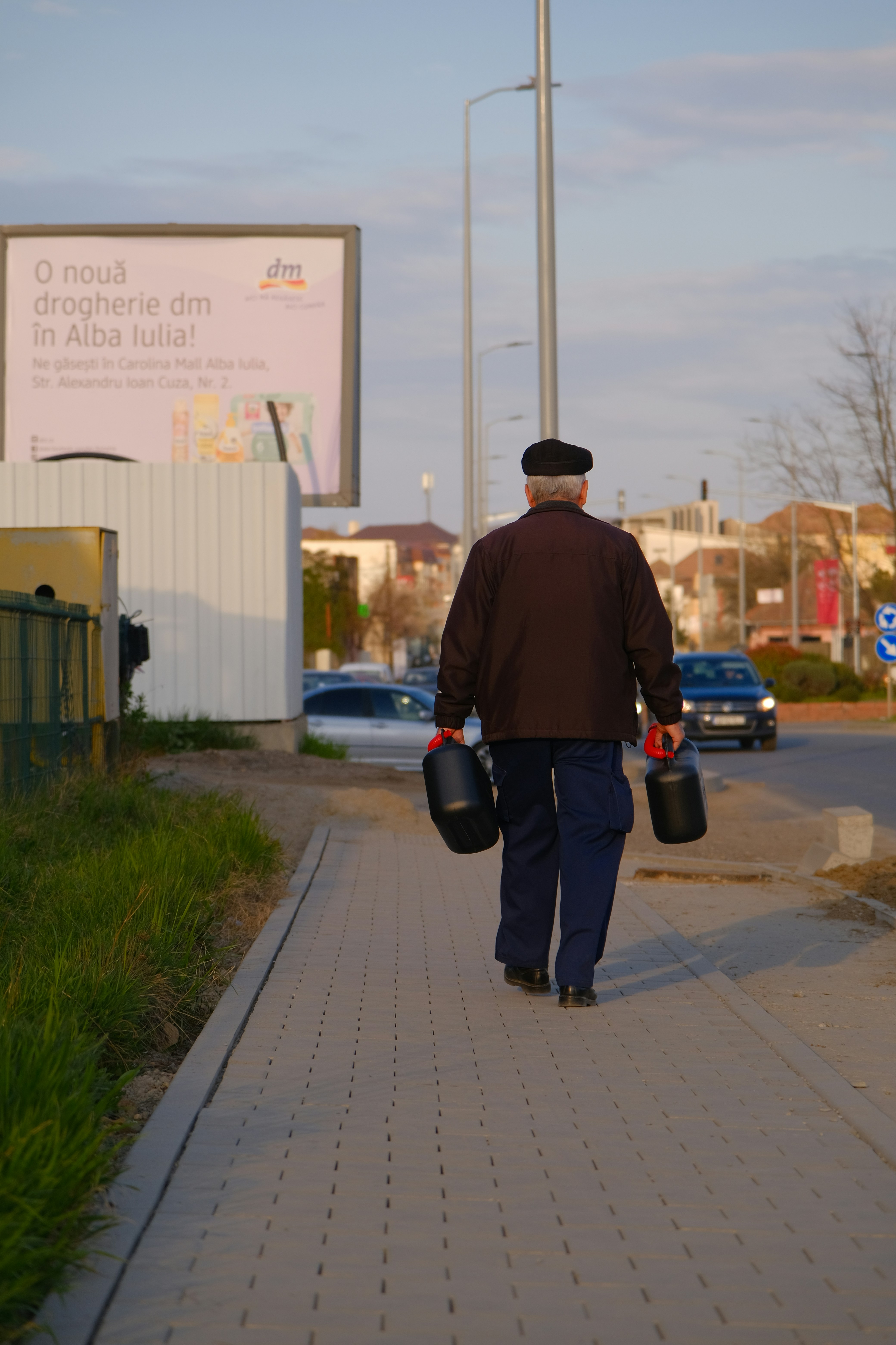 An elderly man walks along a paved sidewalk under the bright daylight, carrying two black containers with red handles. Dressed in casual attire and a hat, he moves away from the camera, towards a bustling urban setting with cars, streetlights, and a billboard advertising a new store in Alba Iulia. The scene captures a moment of everyday life in a modern city, highlighting the simplicity and routine of daily activities.