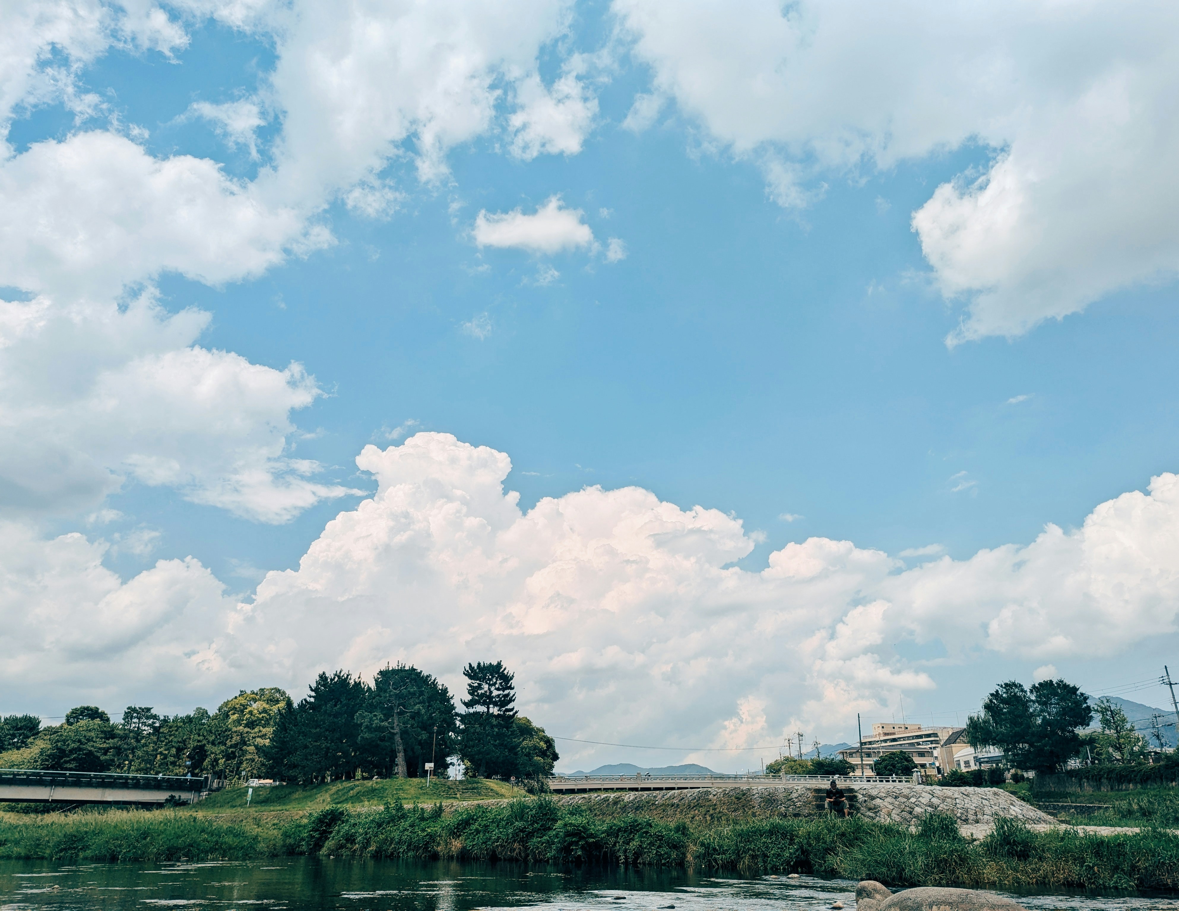 Lush greenery borders a calm river under a vibrant blue sky dotted with fluffy clouds.
