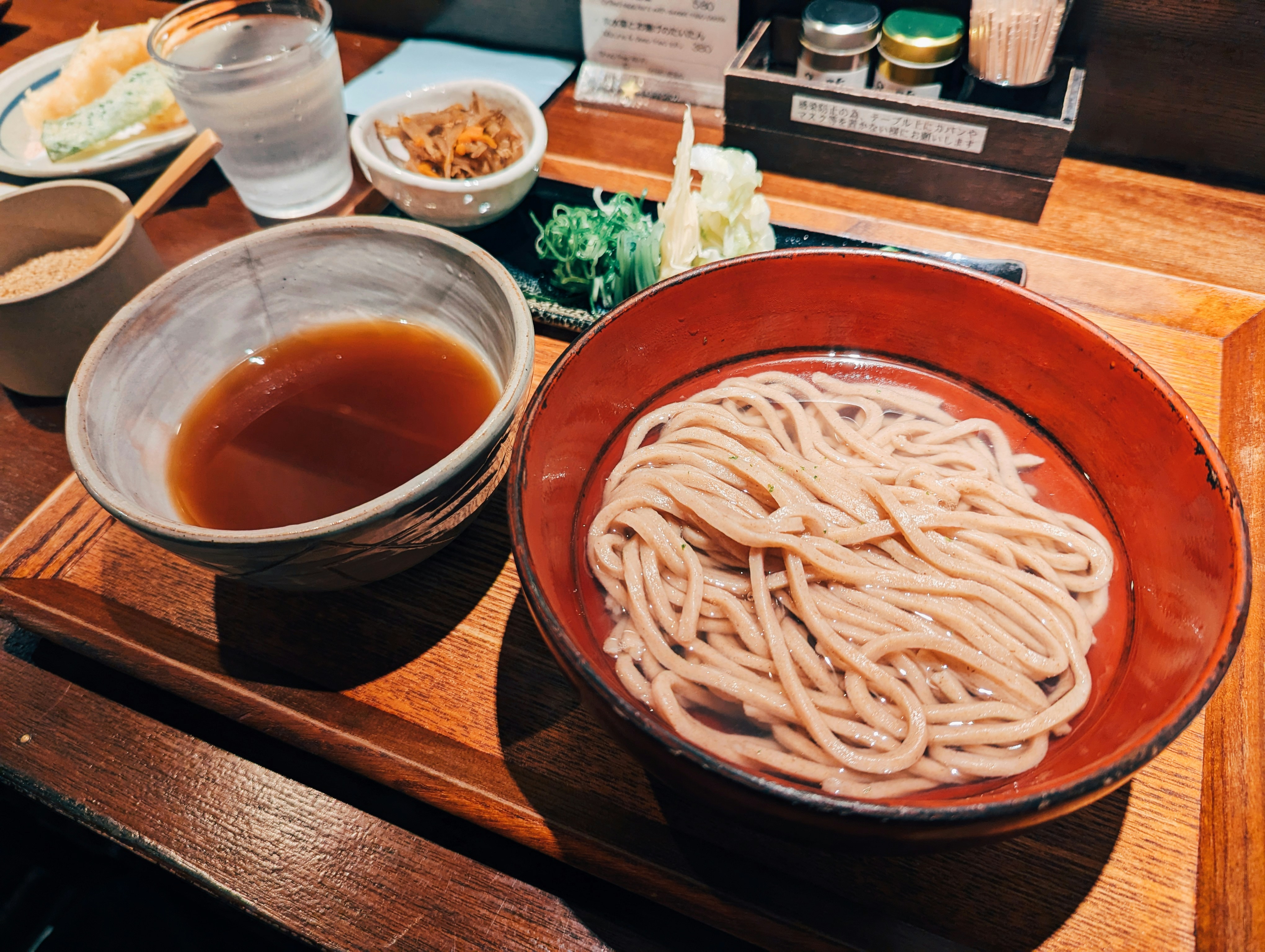 Bowl of udon noodles served with dipping sauce and garnishes on a wooden tray.